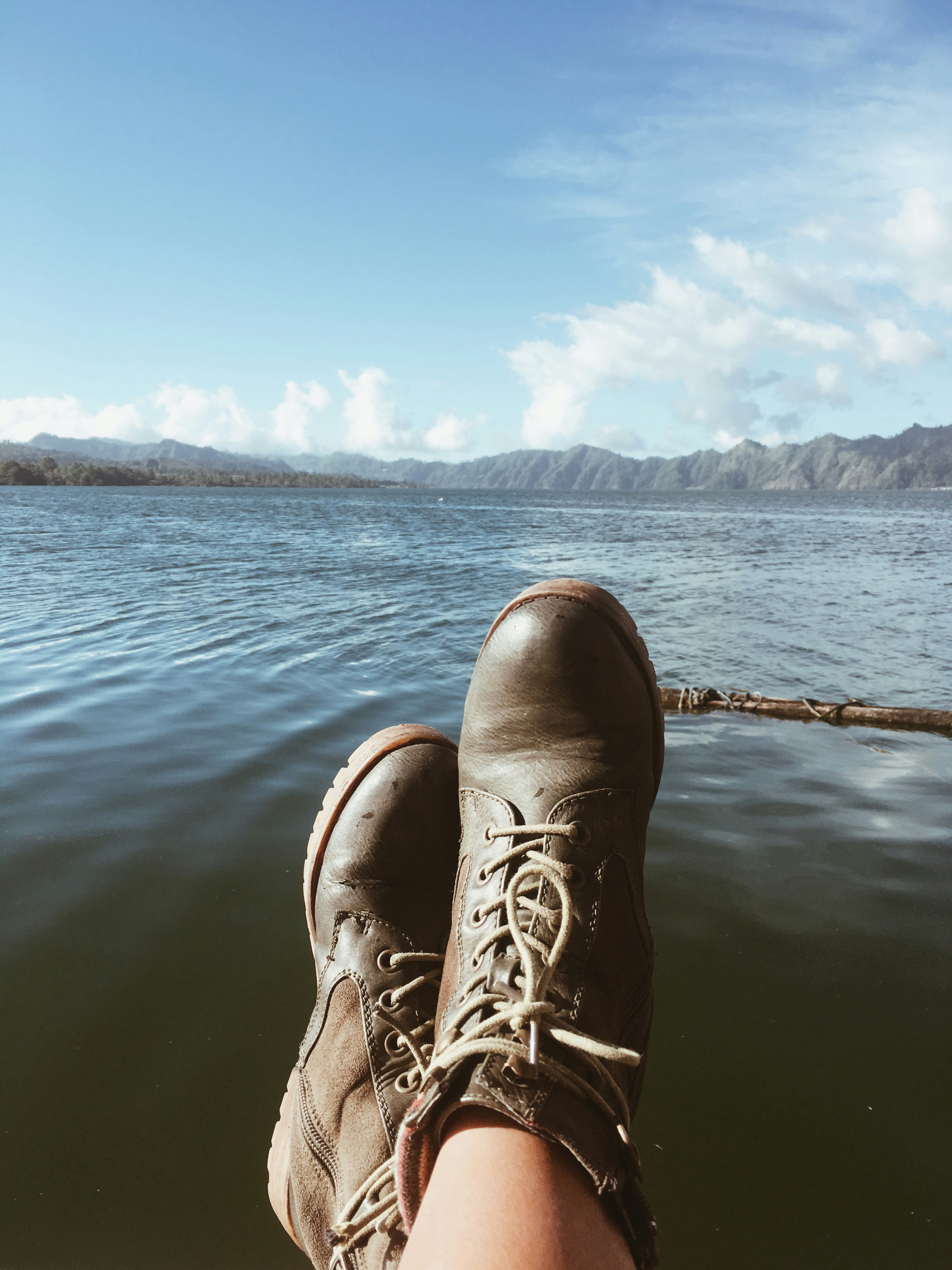 Worn leather boots resting on the edge of a tranquil lake, with mountains and a clear sky in the background.
