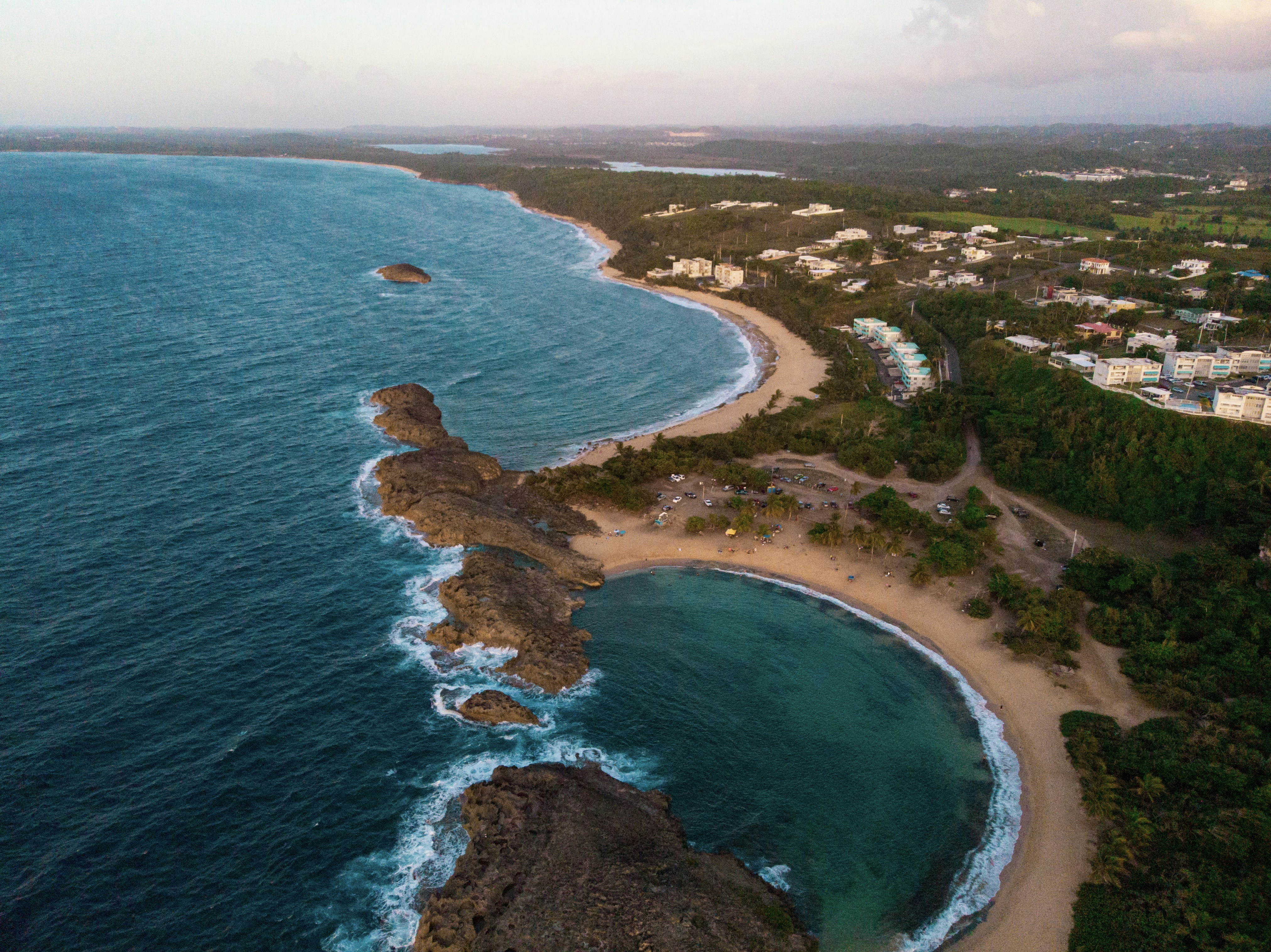 Aerial view of a curved coastline with turquoise water and sandy beach, bordered by lush greenery.