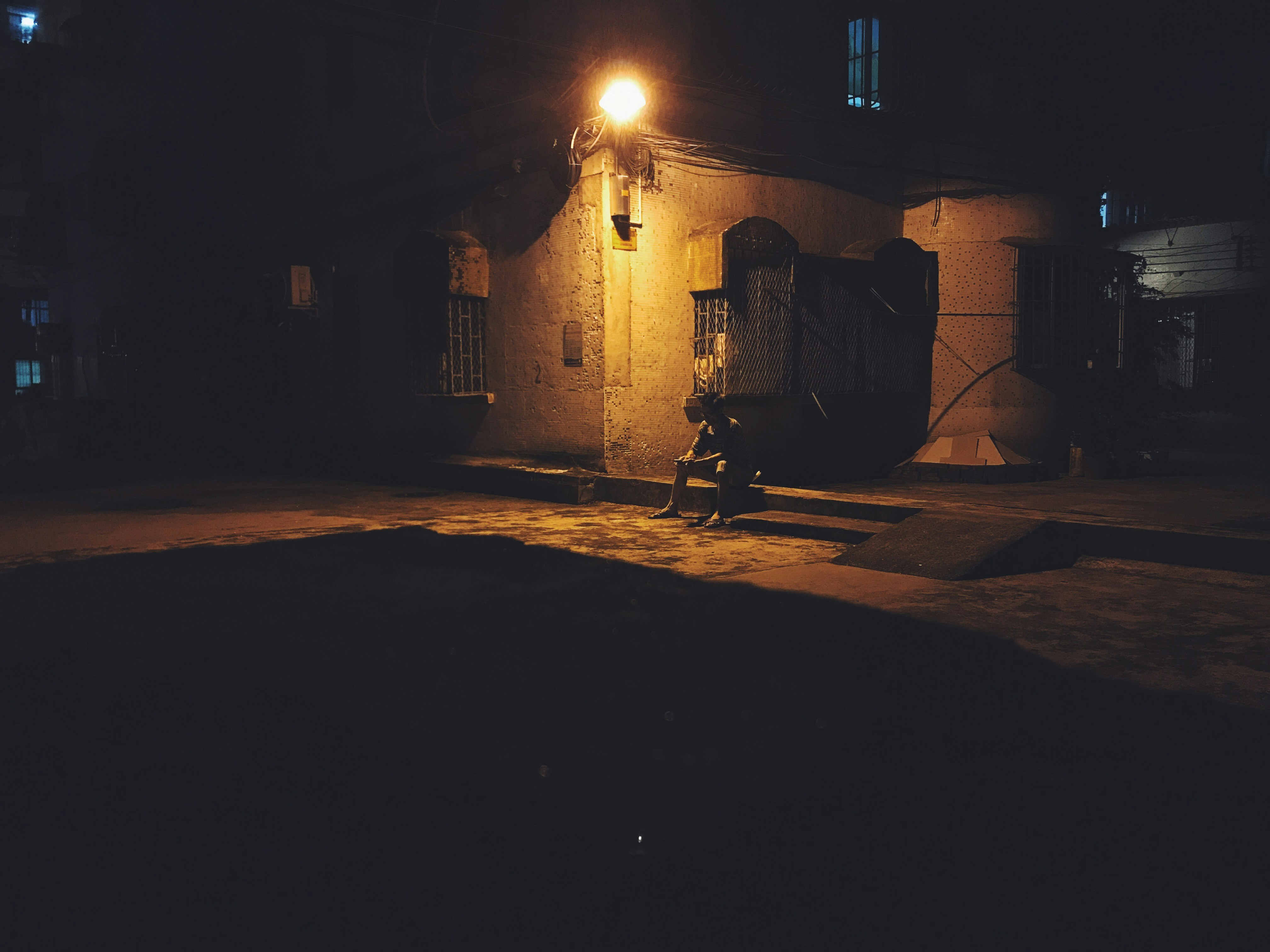A solitary figure sits on a ledge, illuminated by a warm streetlight in a dimly lit urban setting.