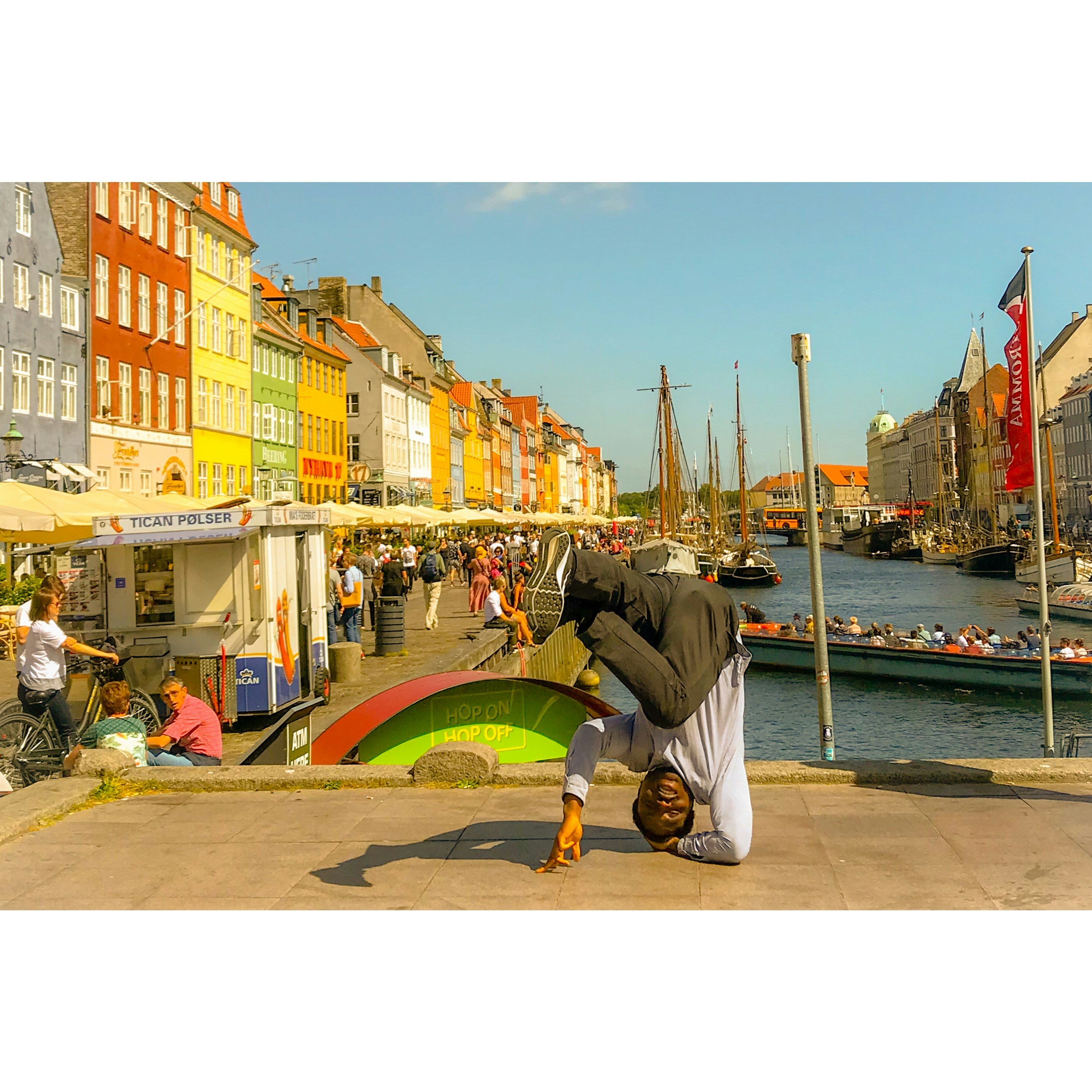 A performer executing a handstand on a bustling waterfront promenade lined with colorful buildings and boats. The lively atmosphere captures the essence of street performance culture.