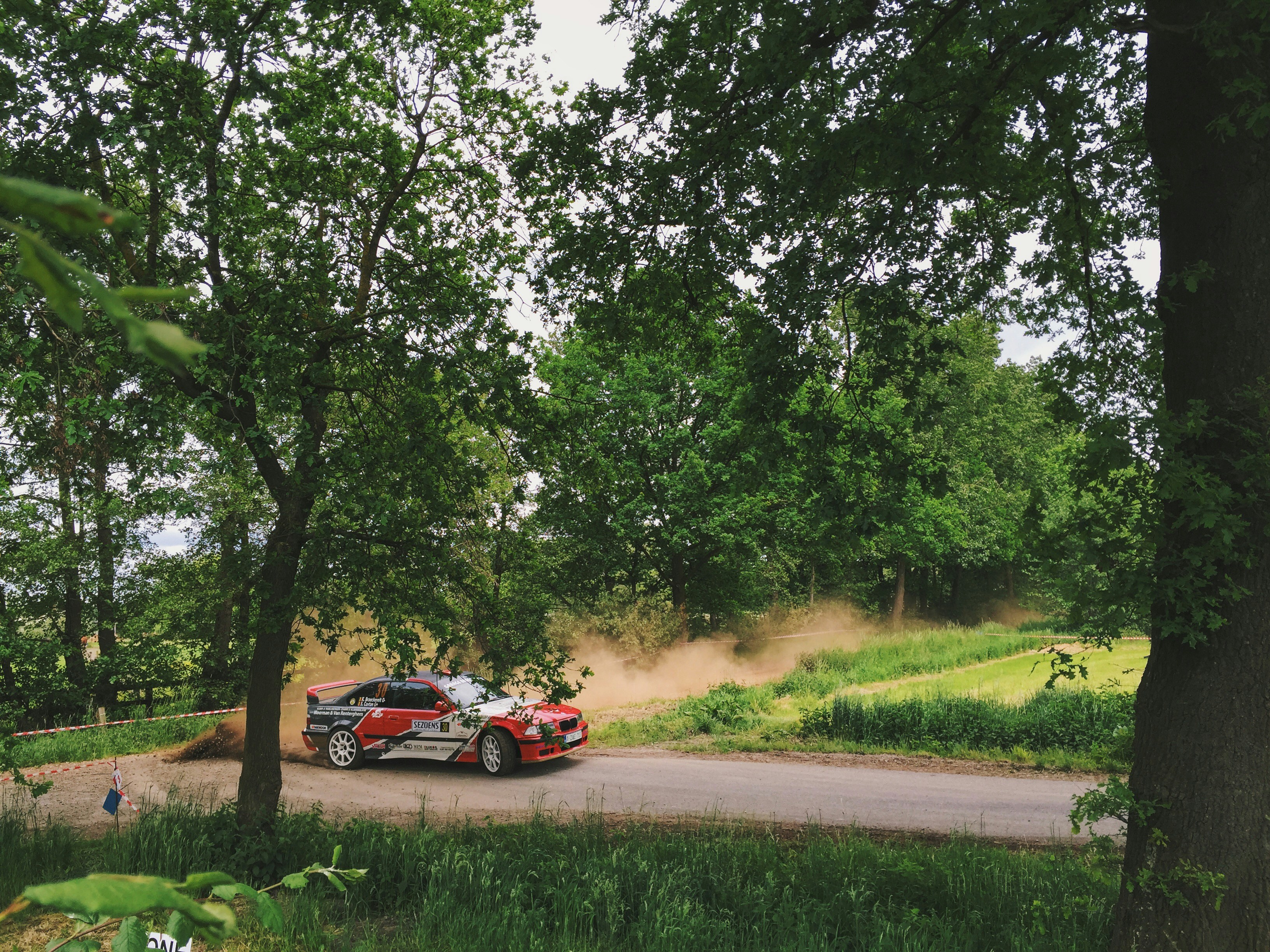 Red and white sedan speeds along a tree-lined road, kicking up dust.