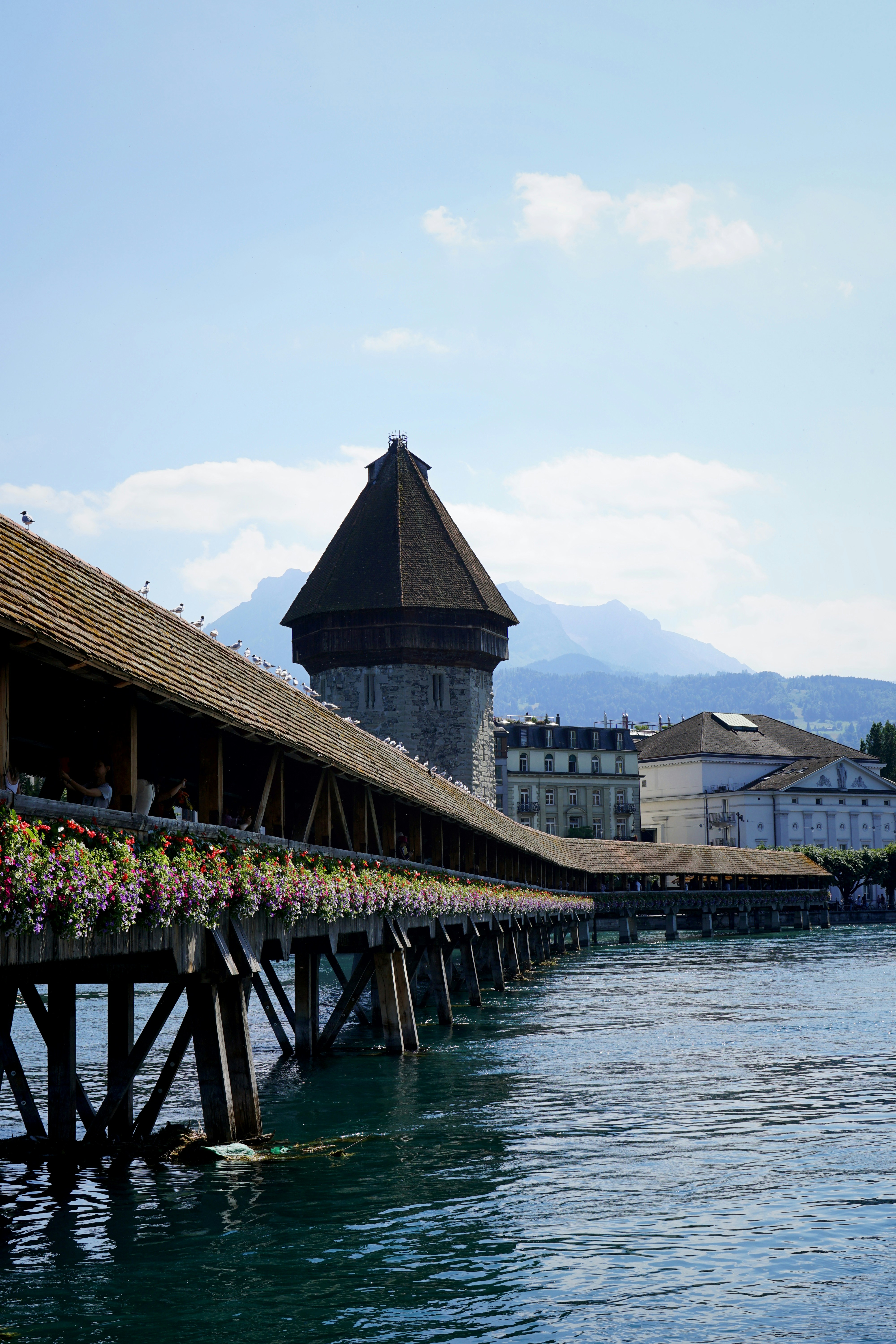Historic wooden bridge adorned with flowers stretches across a serene lake, with a stone tower and mountains in the background.