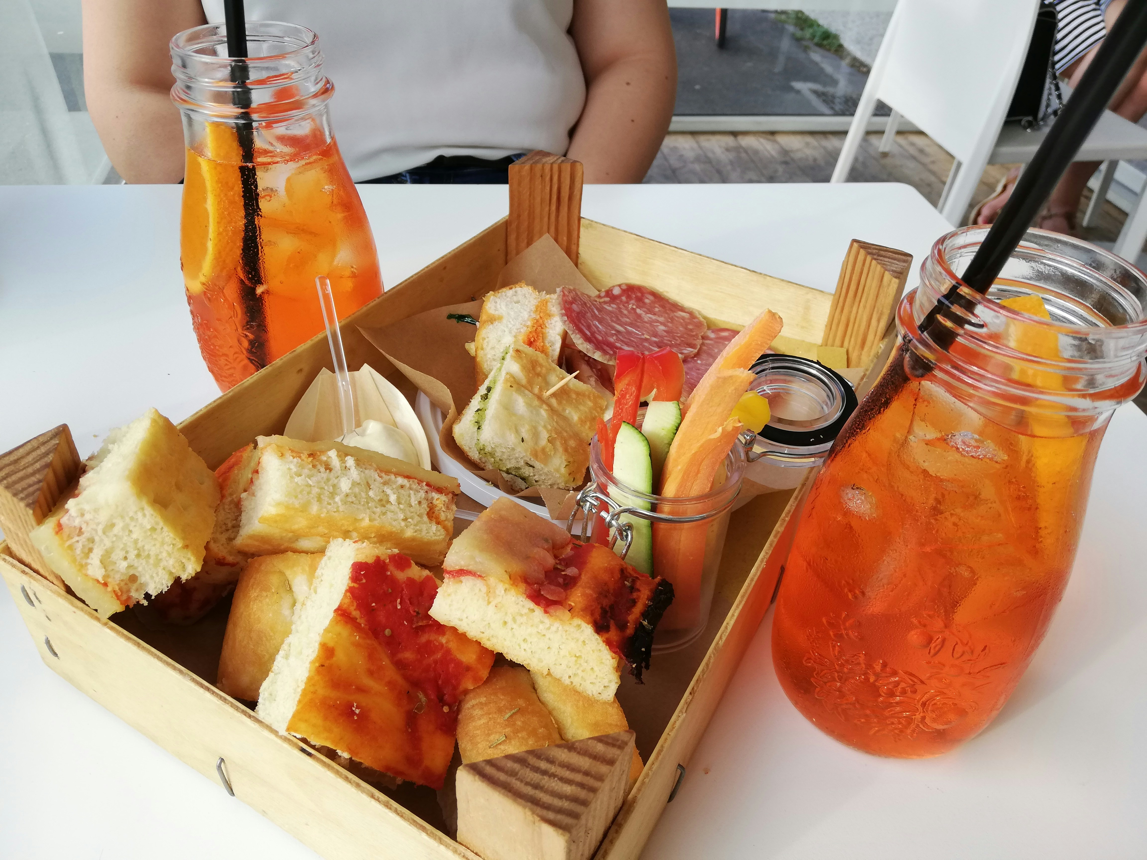 A wooden tray filled with a variety of sandwiches, breads, and fresh vegetables, accompanied by two refreshing drinks in mason jars.