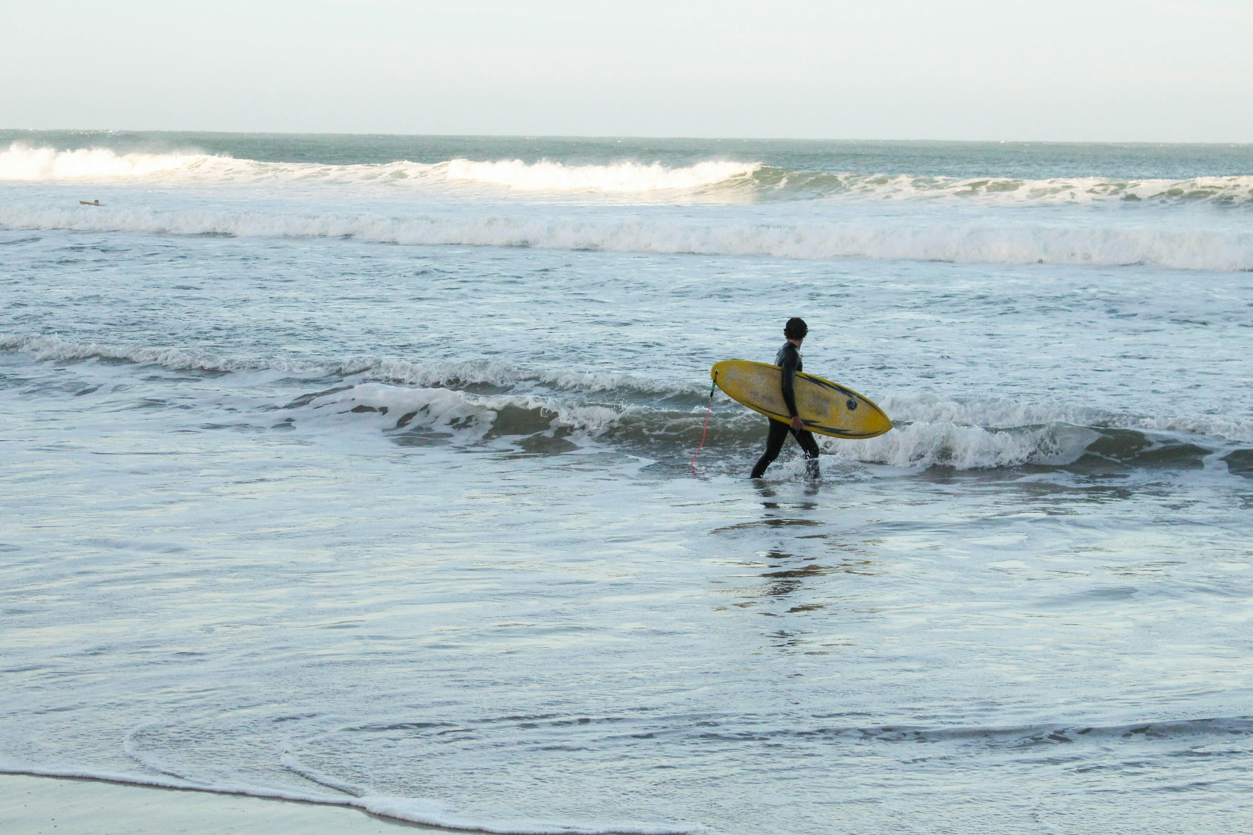 Surfer with a board walking towards gentle ocean waves under a clear sky.