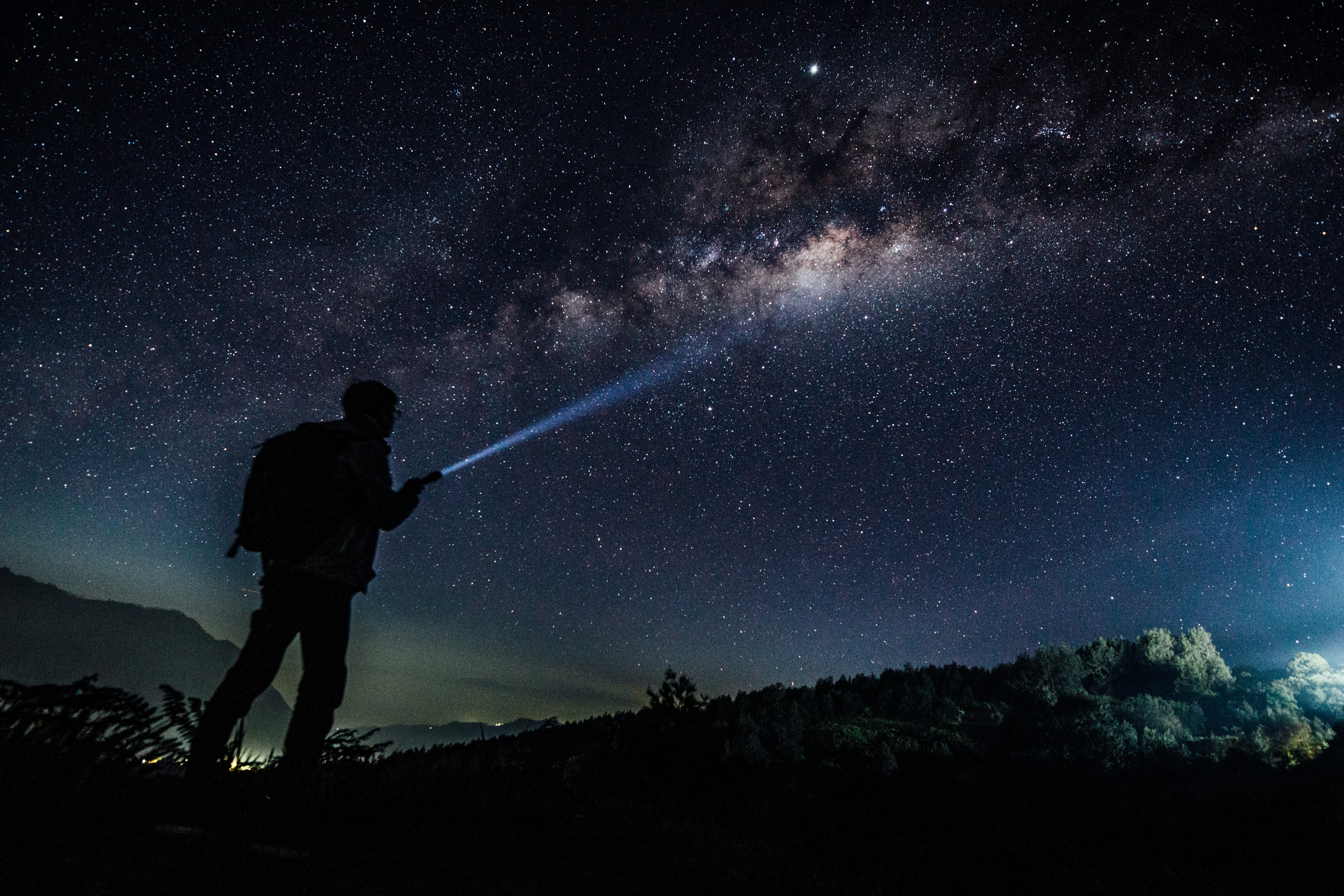 Silhouette of a hiker pointing a flashlight towards the starry sky, revealing the Milky Way's brilliance above a serene landscape.