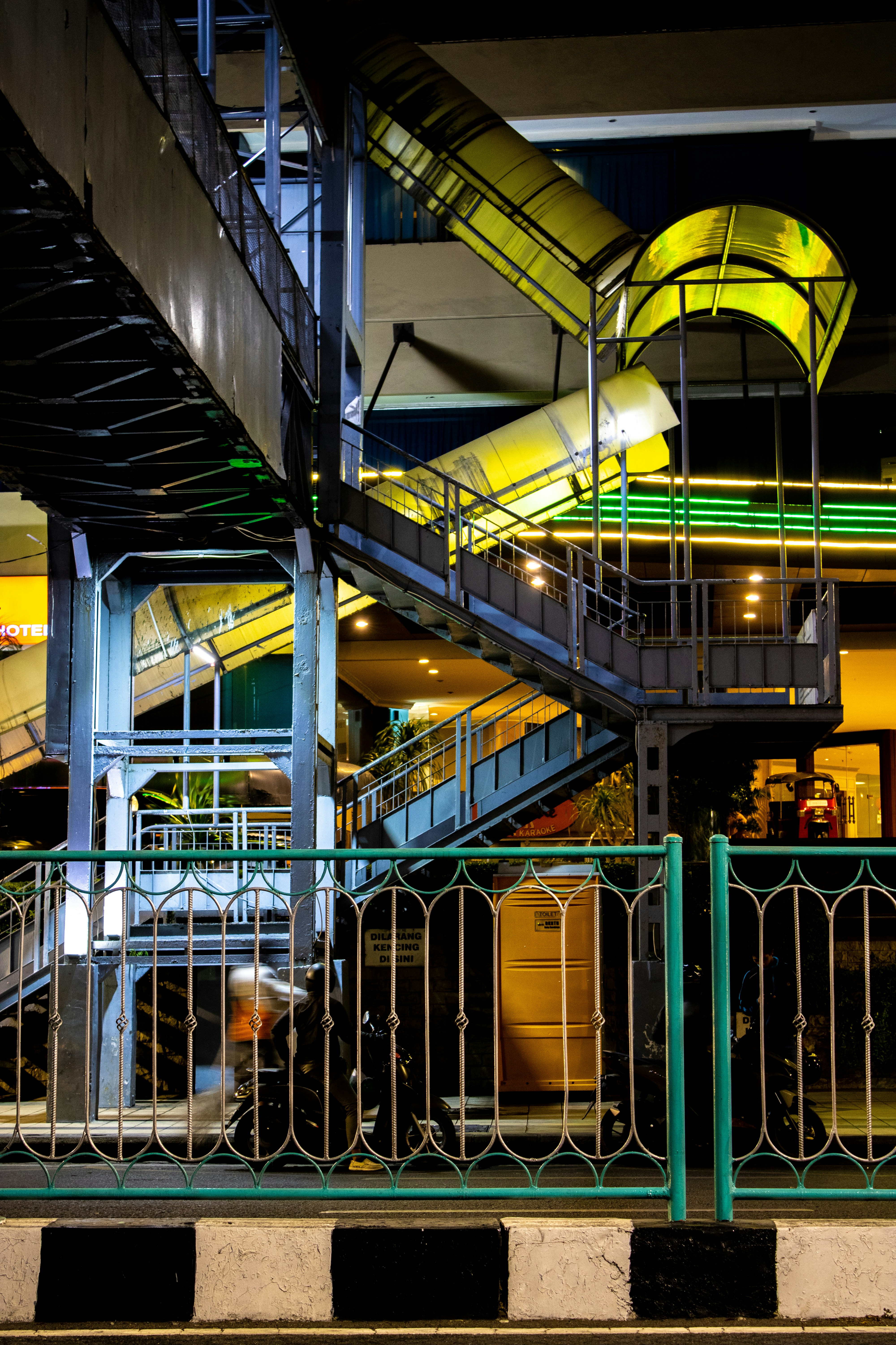 A vibrant pedestrian bridge with illuminated slides and staircases in a bustling urban setting, showcasing modern infrastructure. The scene captures the dynamic interplay of light and structure.
