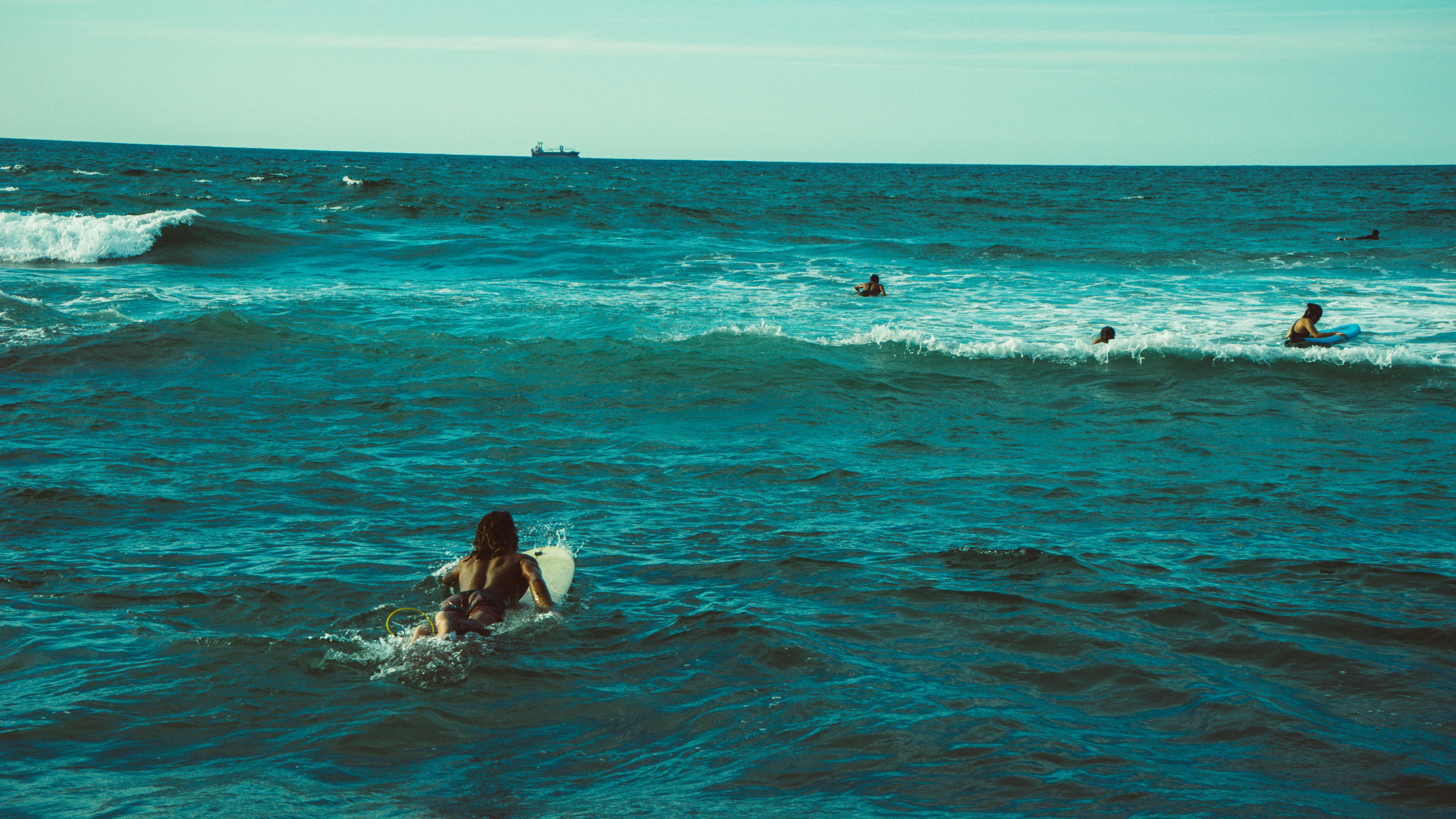 Surfer paddling through azure waters, surrounded by fellow surfers and a distant cargo ship on the horizon.