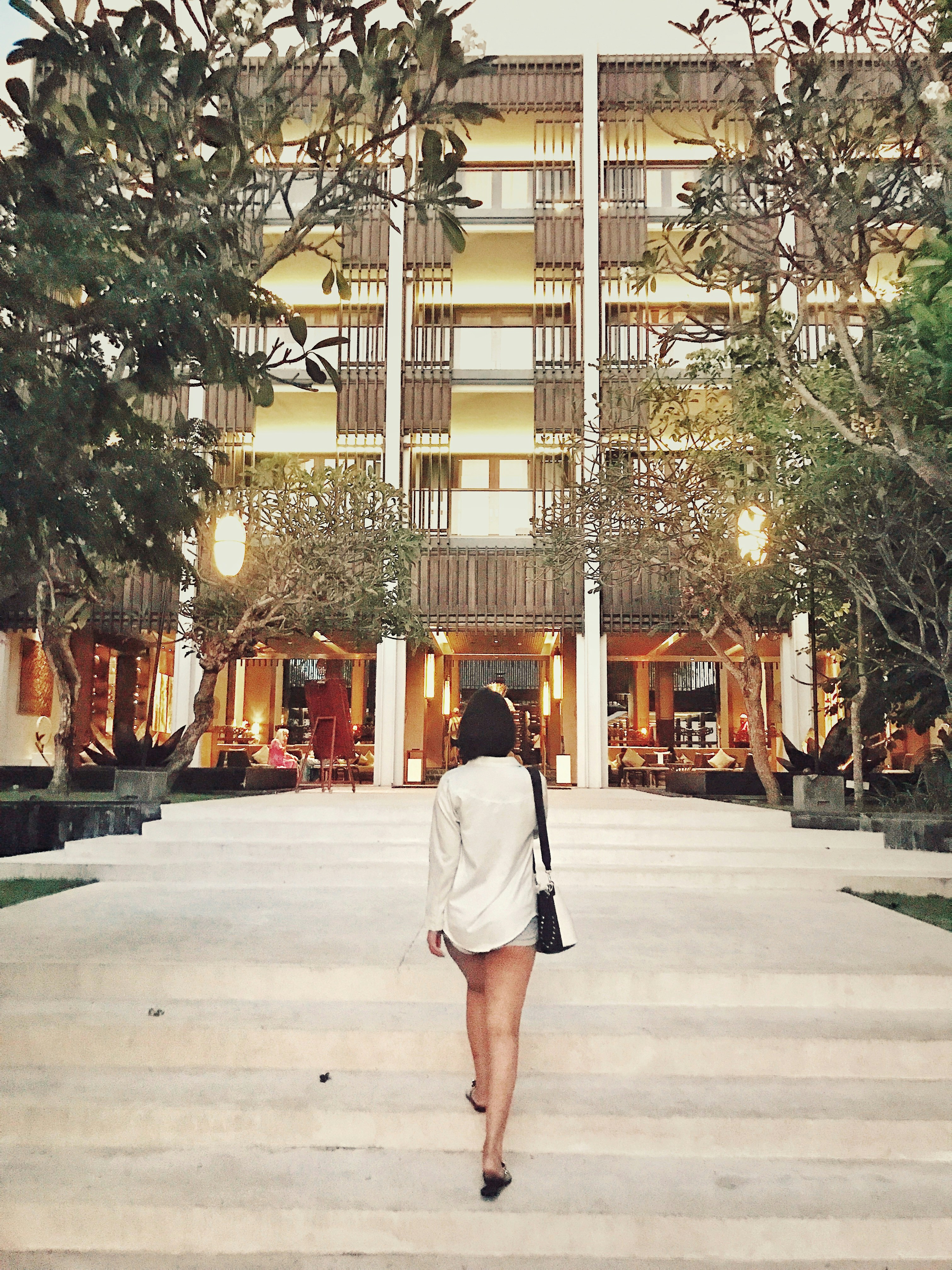 Woman in white shirt walking up steps toward a warmly lit building surrounded by trees.