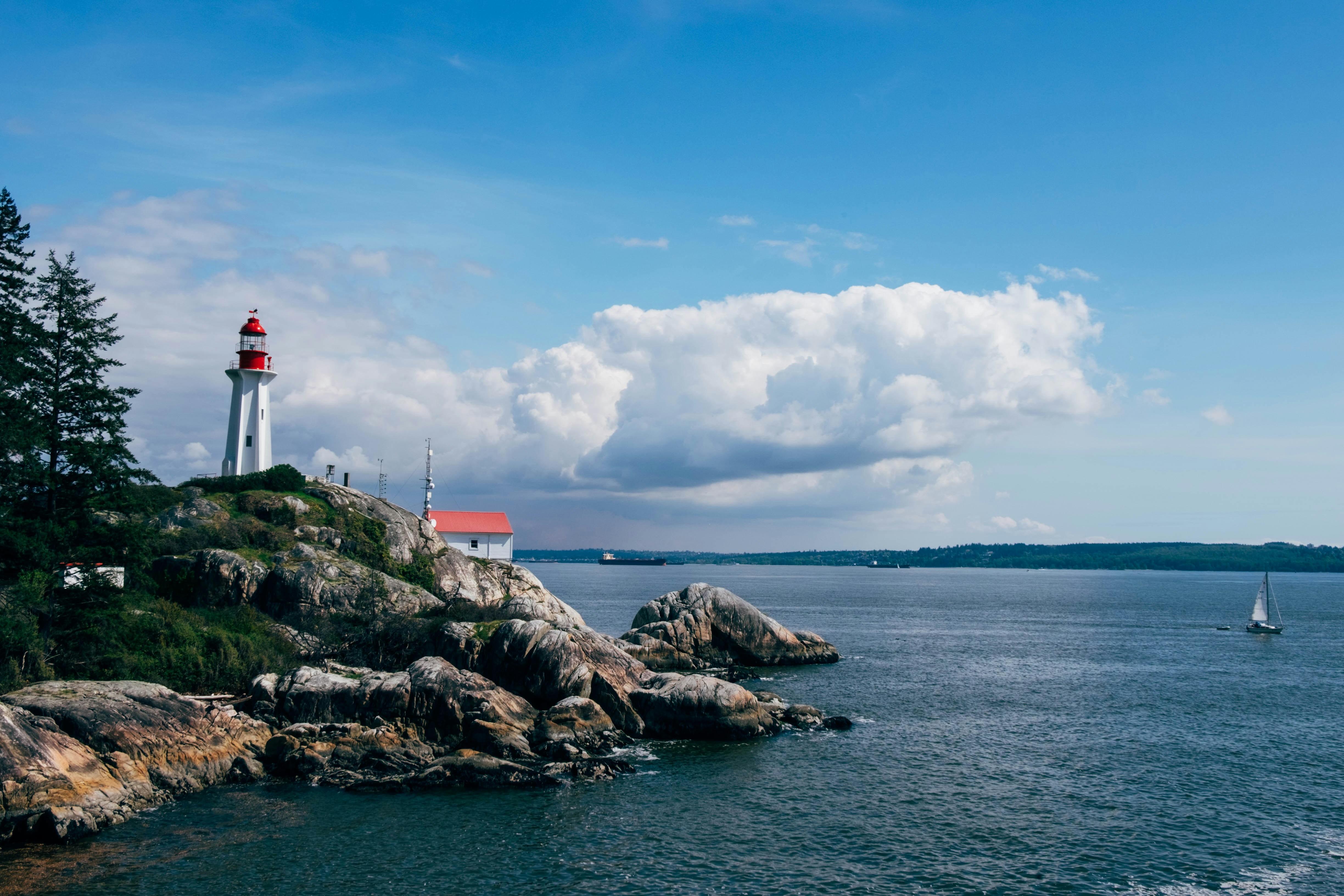 Historic lighthouse standing tall on rocky shores, overlooking calm waters and distant sailboats under a vast sky.