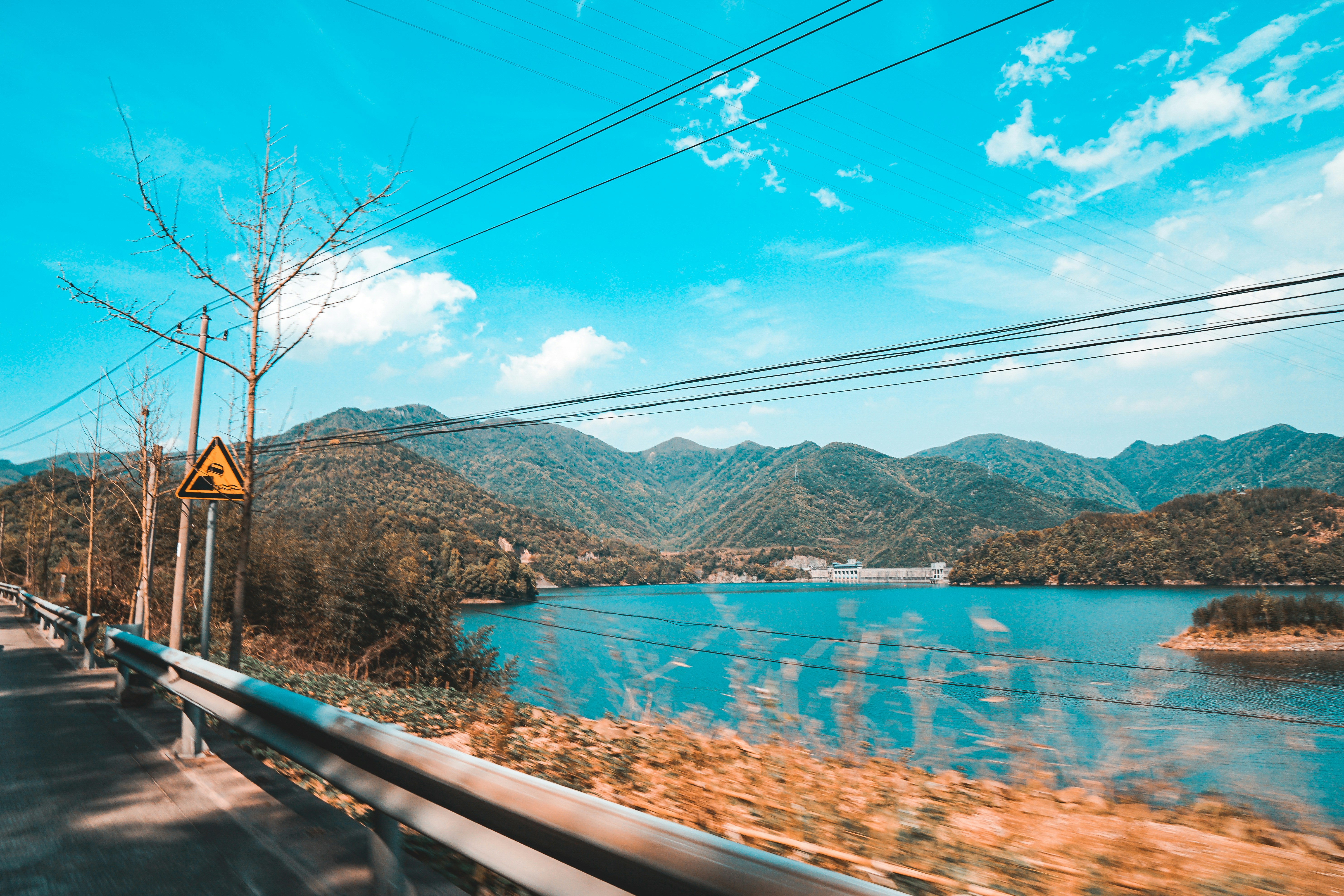 A tranquil lakeside scene with rolling hills and a clear blue sky, punctuated by a caution sign along the roadside. The landscape captures the essence of peaceful exploration.