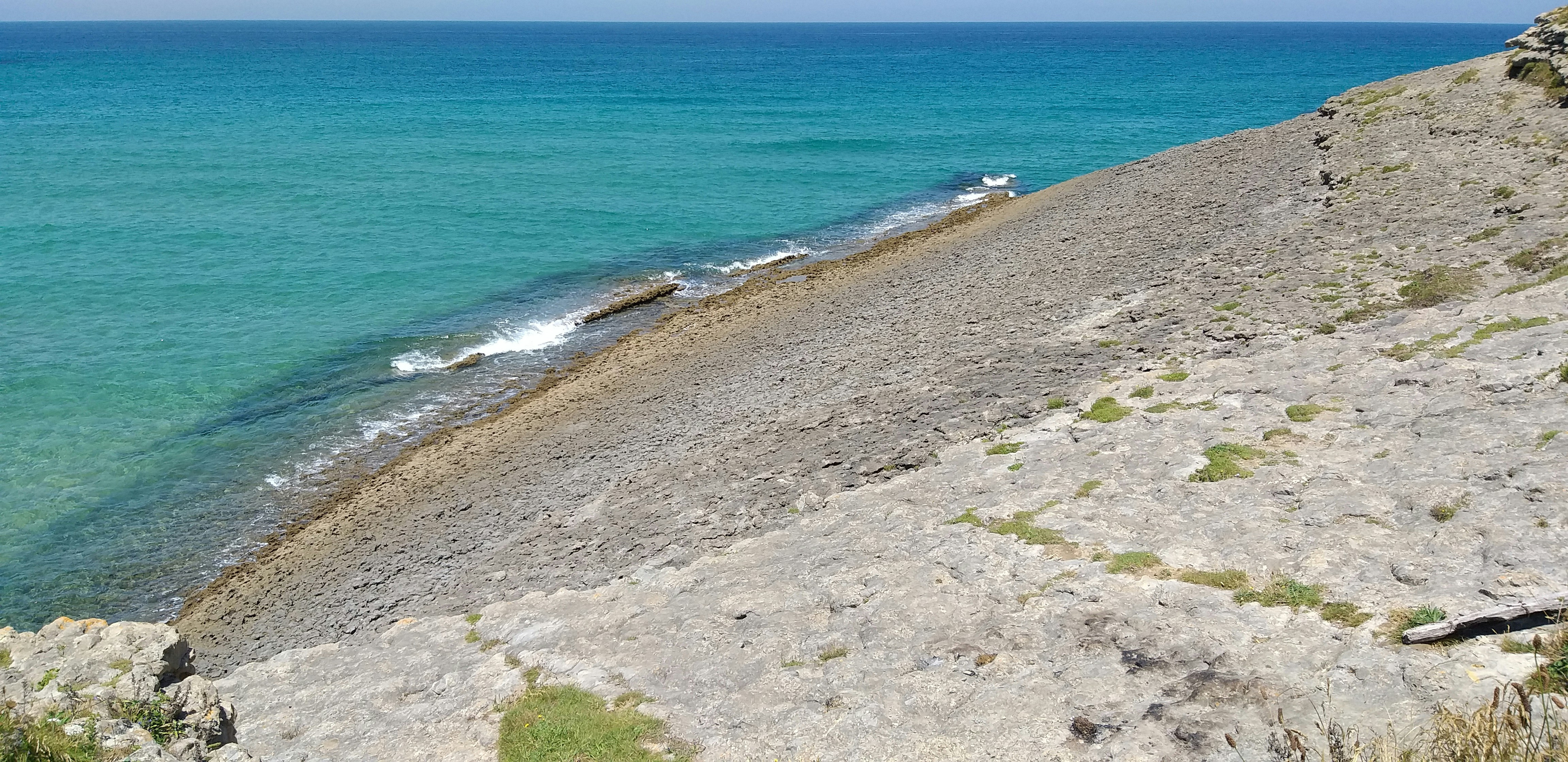Turquoise waves gently lap against a rocky beach with steep cliffs under a clear blue sky.