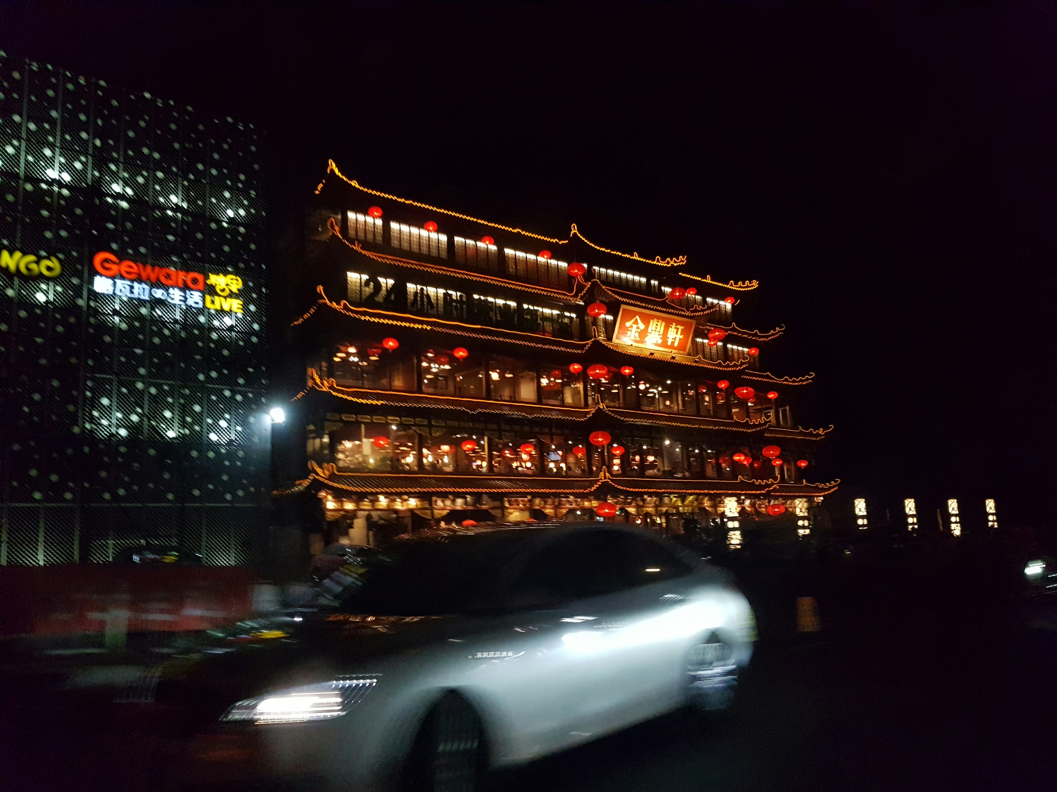 Traditional Chinese architecture adorned with red lanterns stands out against a modern cityscape at night. A passing car adds a sense of dynamic movement.