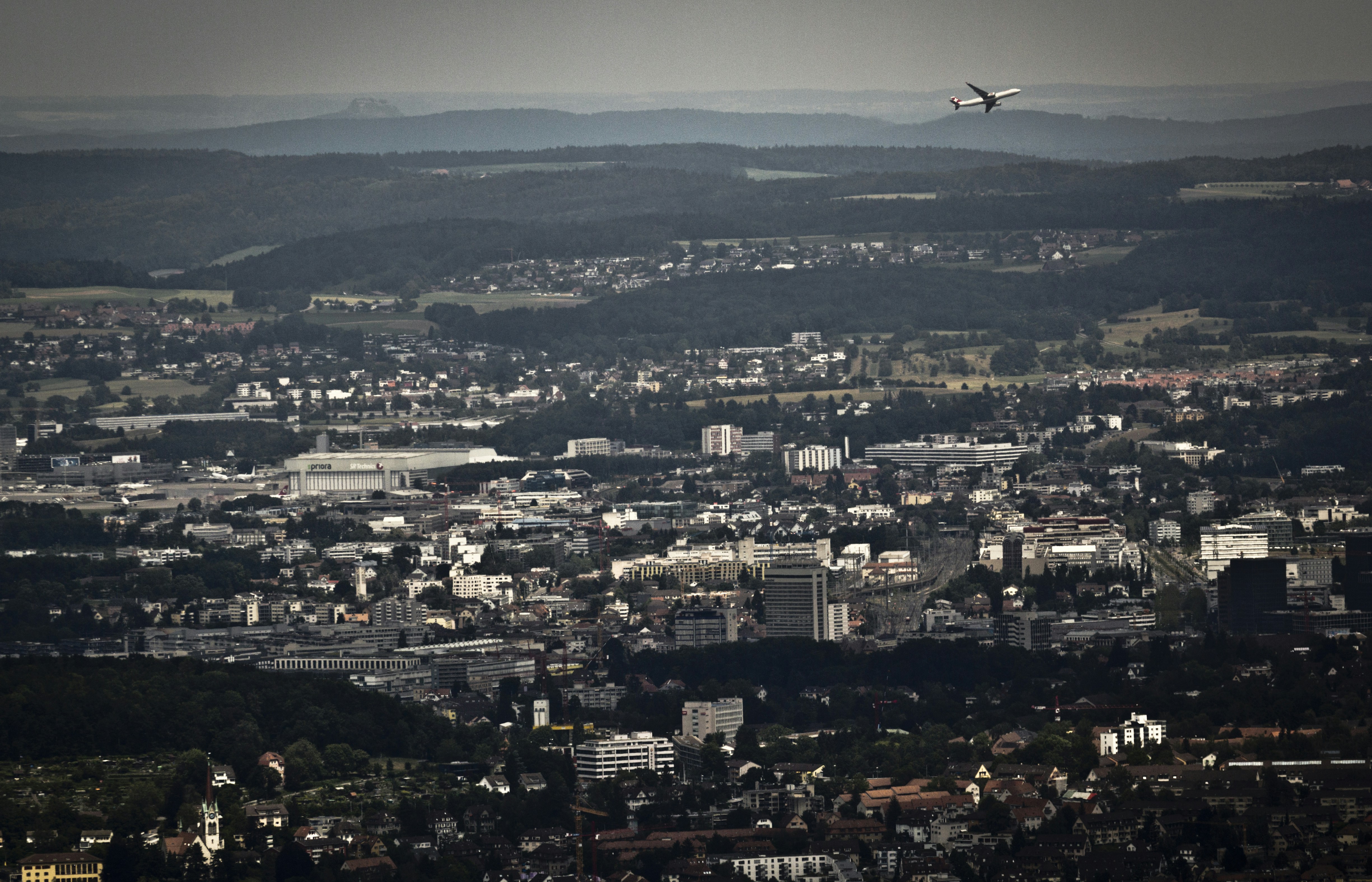 Airplane ascending above Zurich's sprawling cityscape with distant hills under a cloudy sky.
