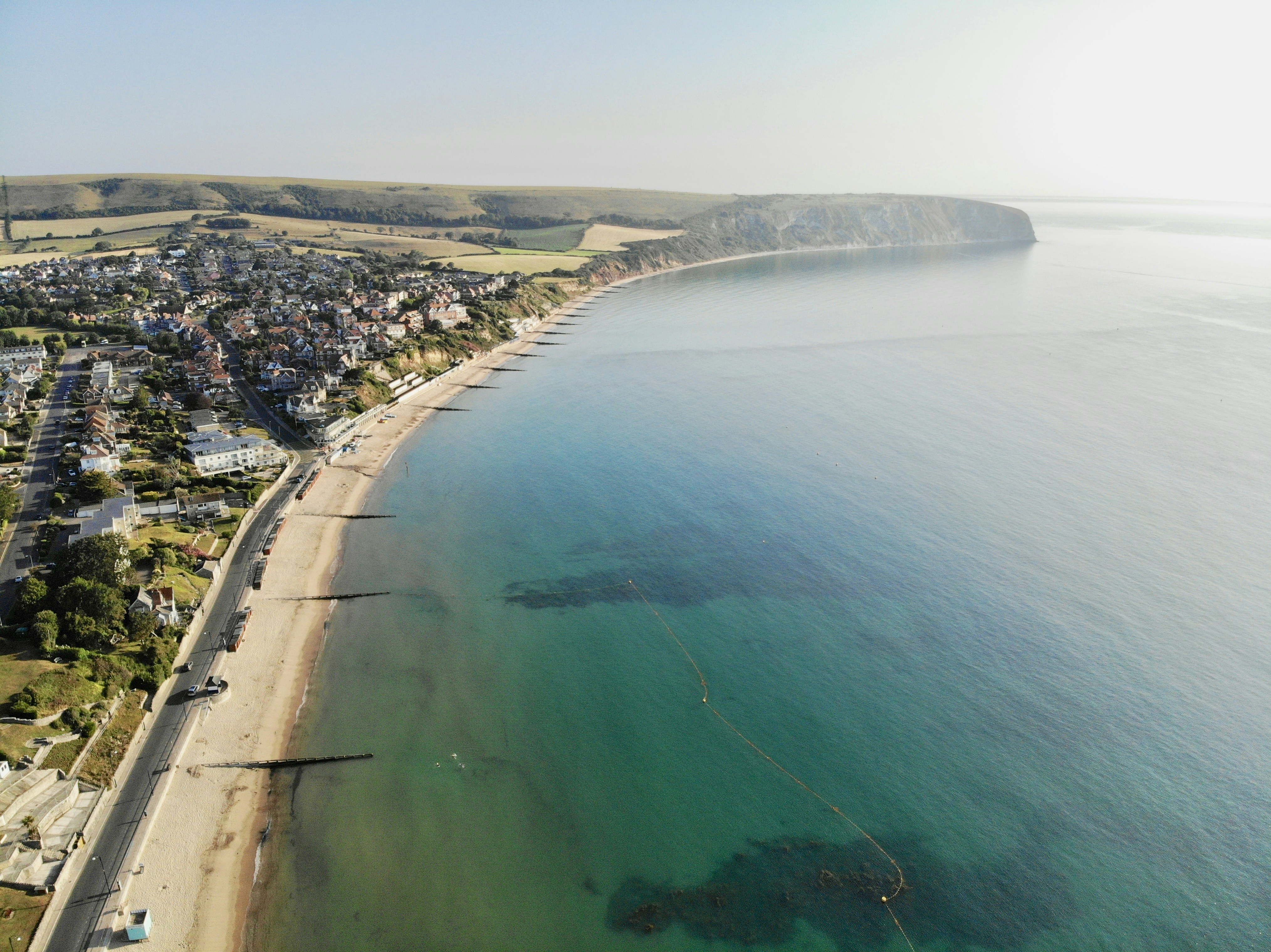 Aerial view of Swanage's coastline with clear turquoise waters and sandy beach curving towards the horizon.