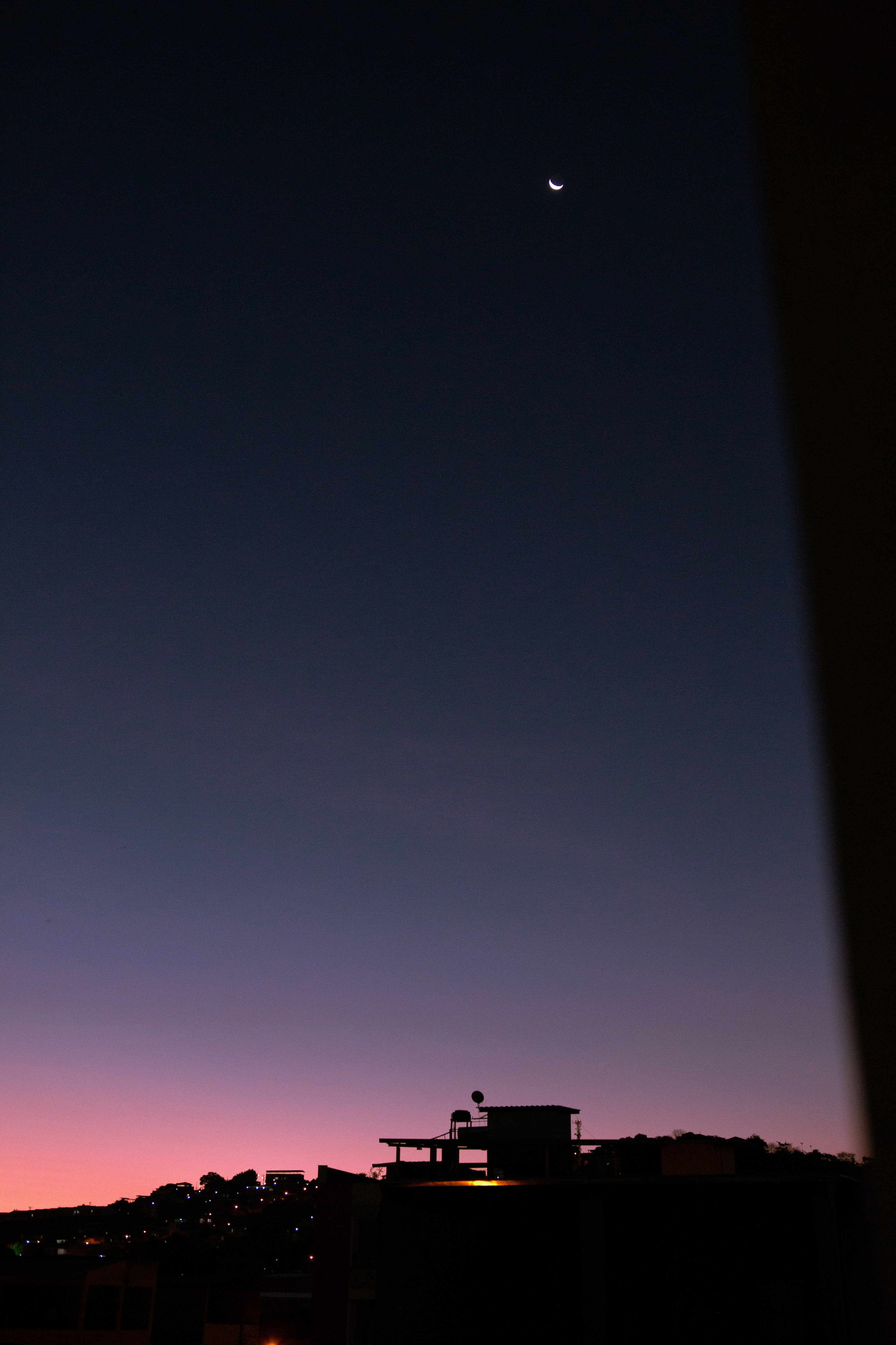 Crescent moon hangs delicately in the twilight sky, silhouetted against a gradient of deep blue and soft pink hues. Rooftops frame the serene scene.