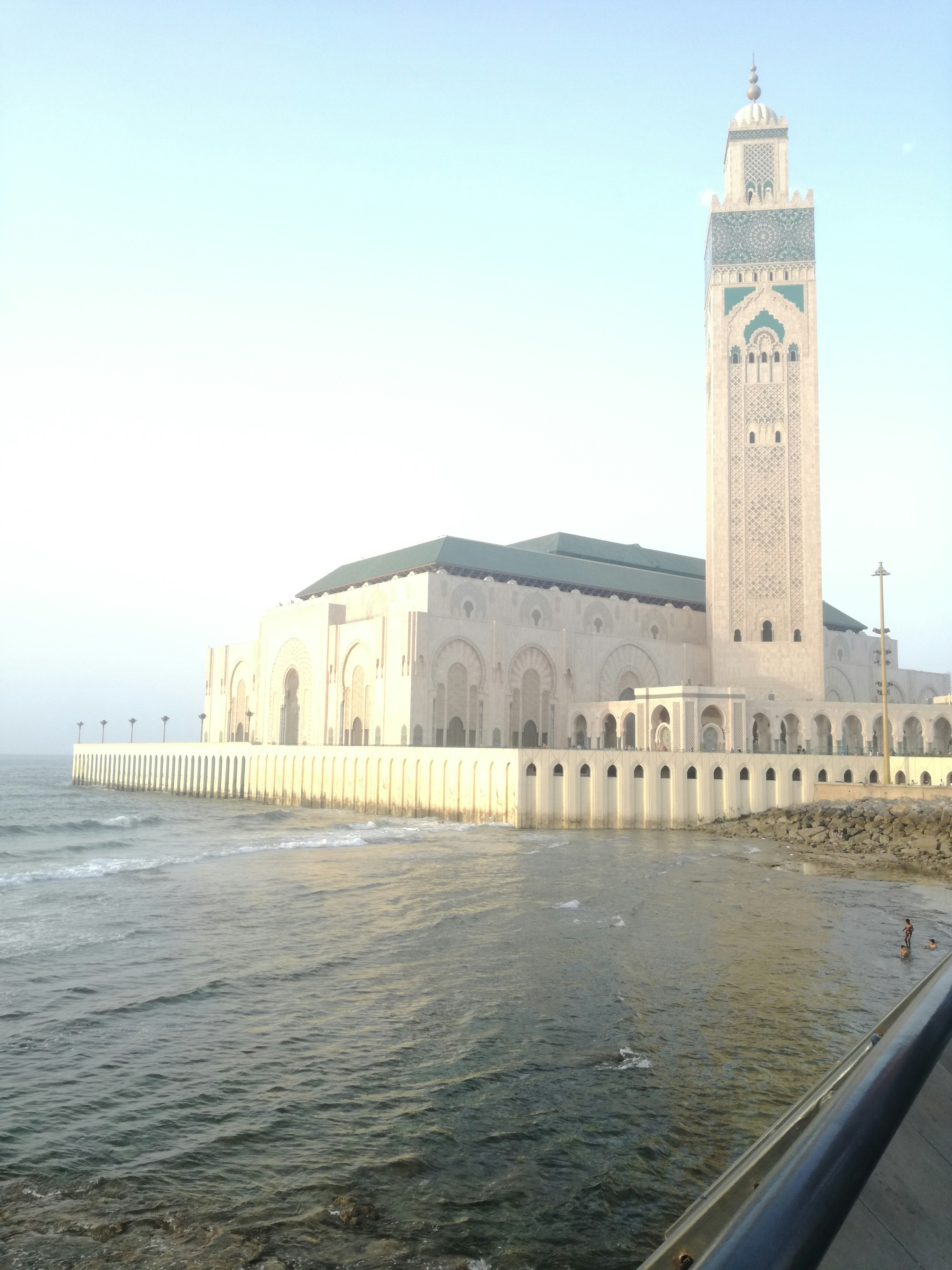 Grand mosque with intricate architecture standing by the ocean, showcasing a towering minaret against a soft sky.