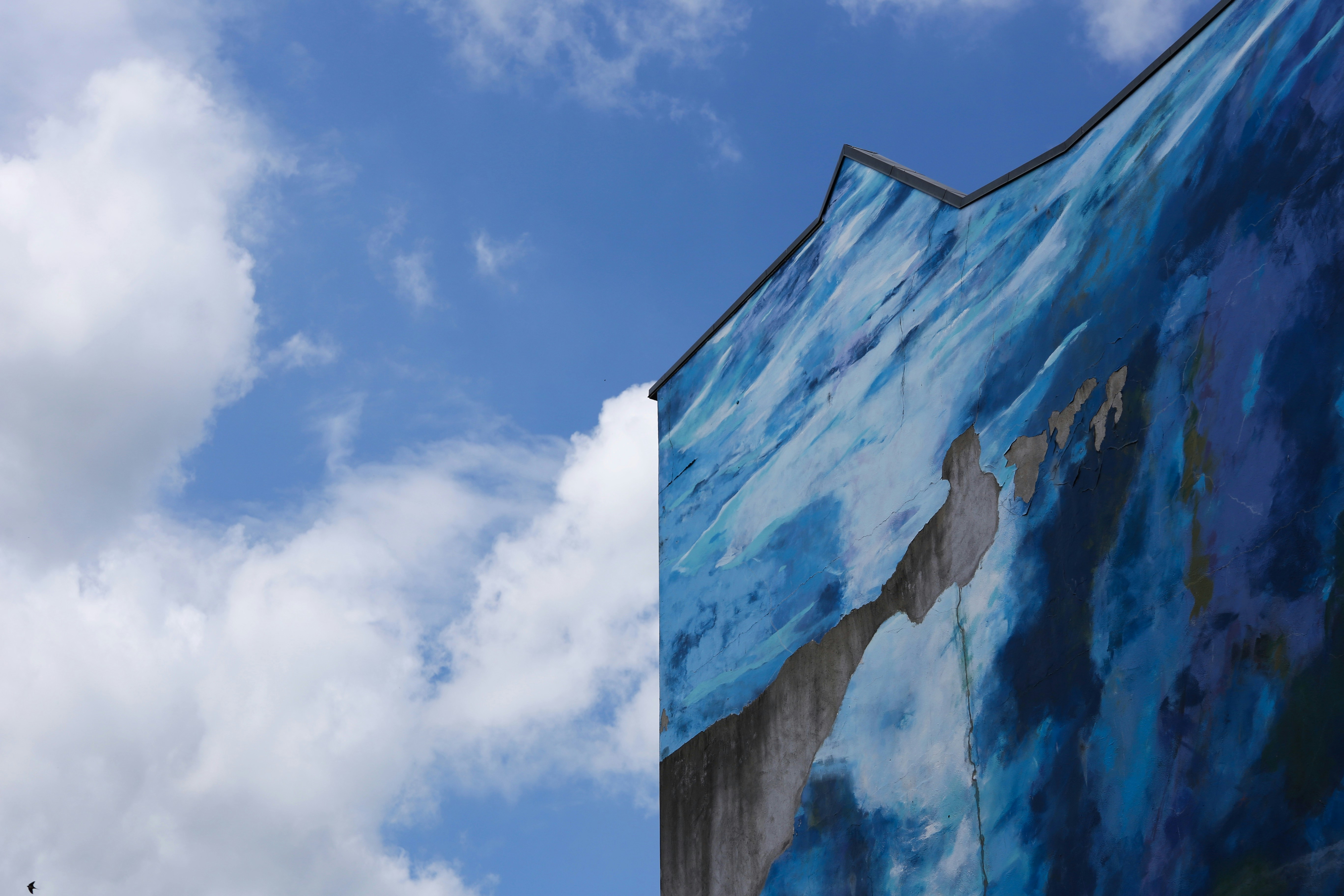 Vibrant mural blending shades of blue against a backdrop of fluffy clouds and azure sky. The artwork captures a moment of artistic expression on a building's facade.