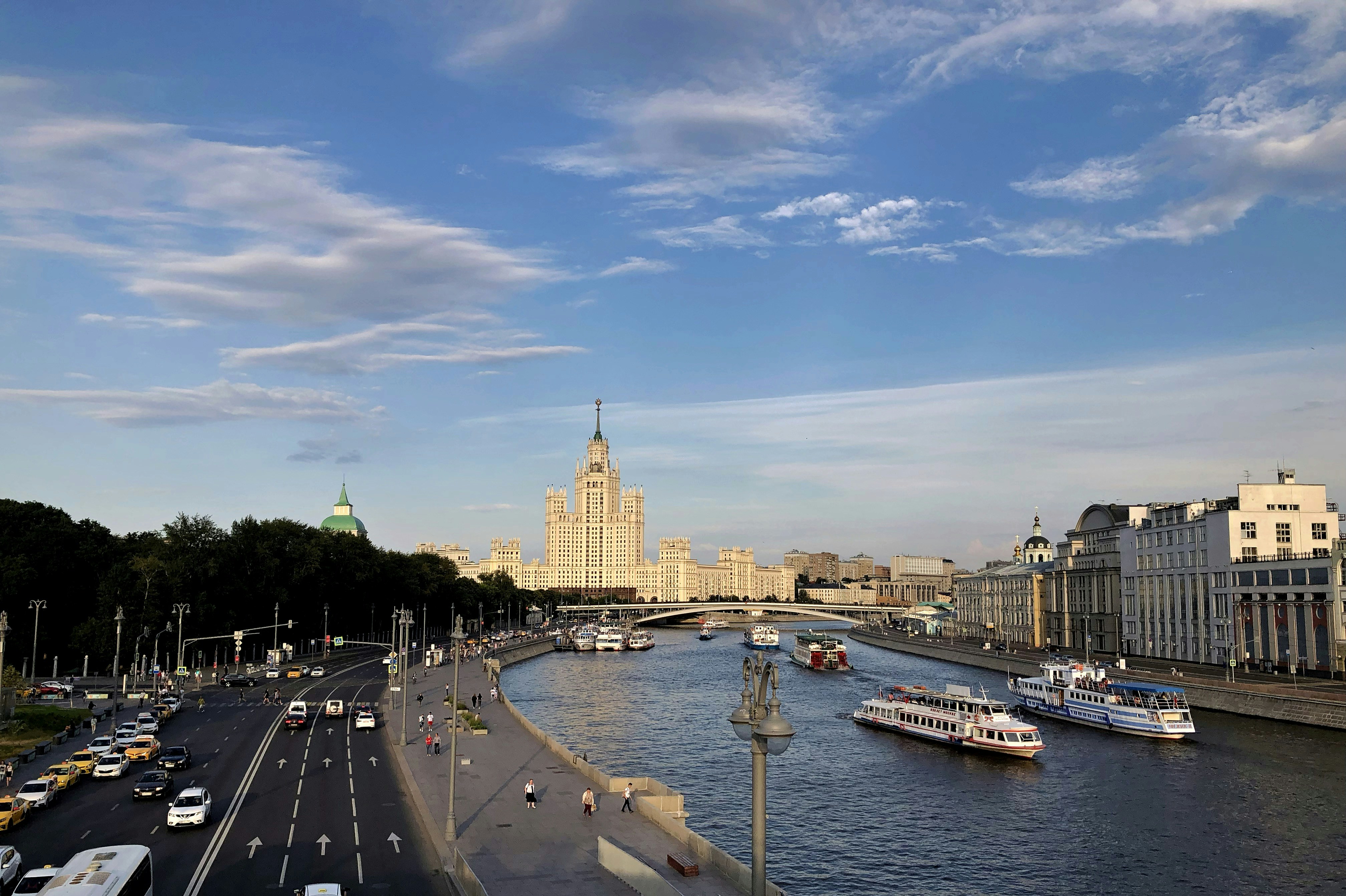 The Moscow River flows gently past the iconic Stalinist skyscraper, framed by a vibrant sky and bustling city life. Boats glide along the waterway, adding to the scene's dynamic energy.