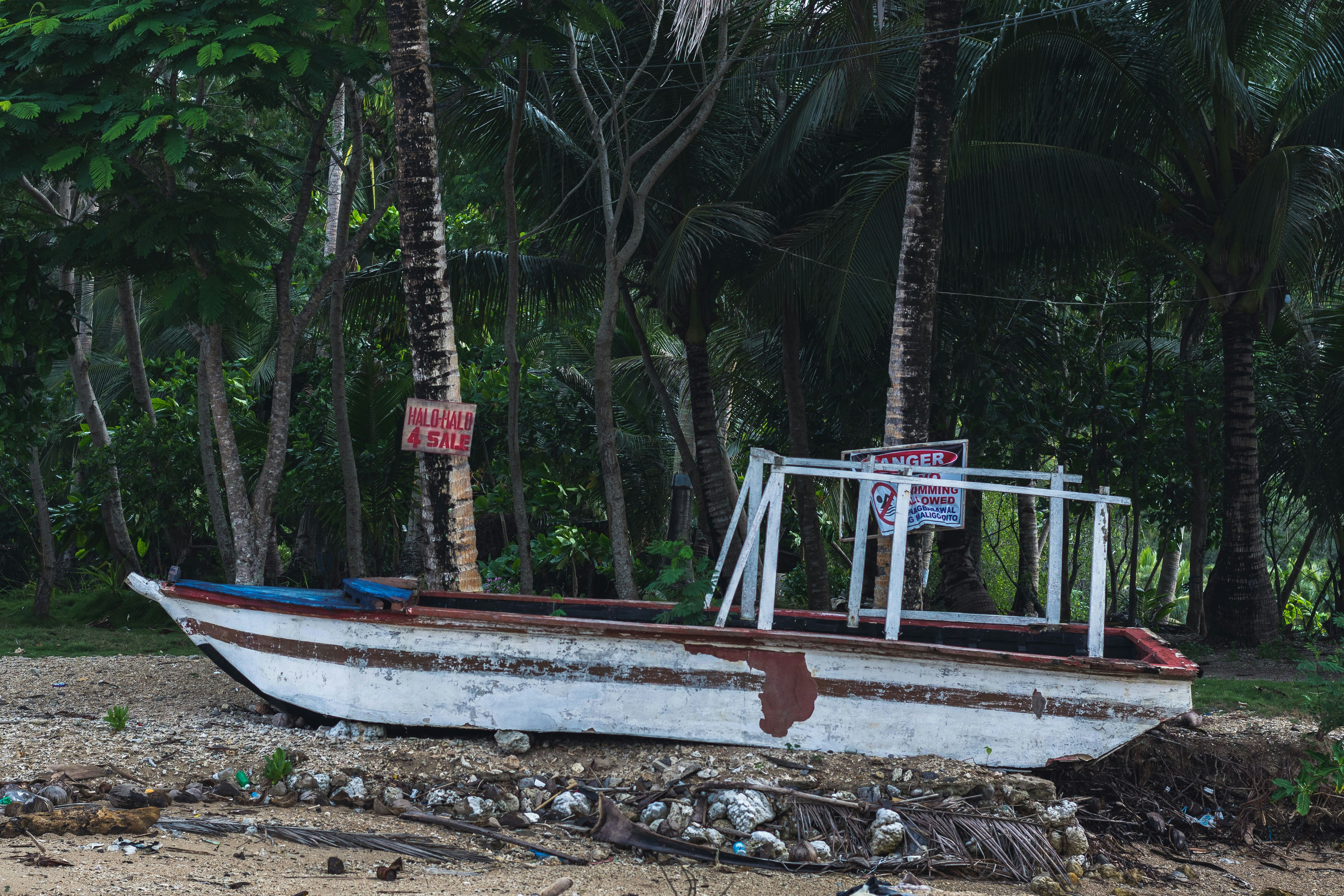 Abandoned fishing boat resting on a sandy beach, surrounded by lush greenery and palm trees. Signs indicate the area is restricted.
