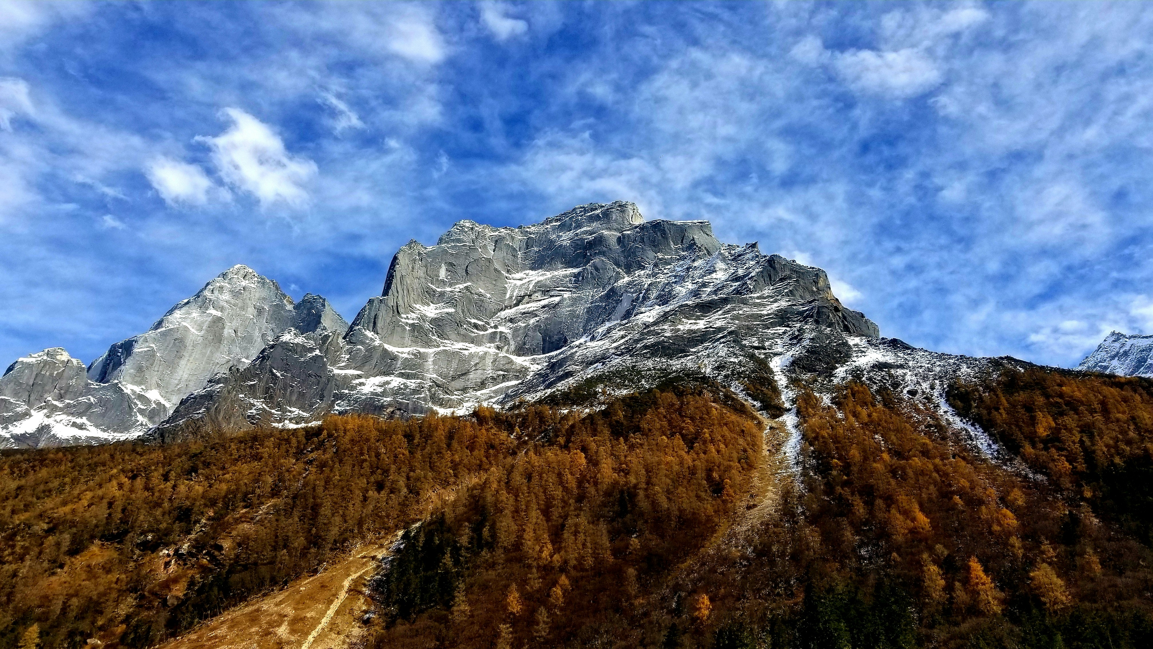 Snow-dusted mountain rises above vibrant autumn forest under a textured sky.