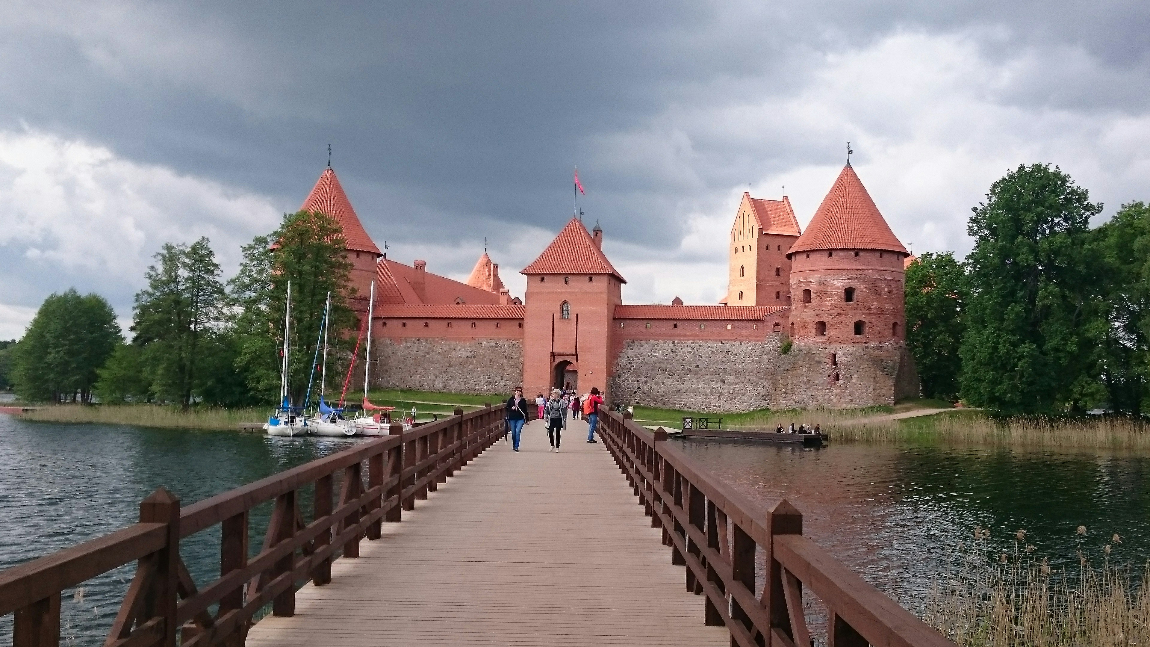 Photograph of a medieval fortress with red cone-topped towers connected by a wooden footbridge over calm water. People walk toward the gate beneath a cloudy sky.