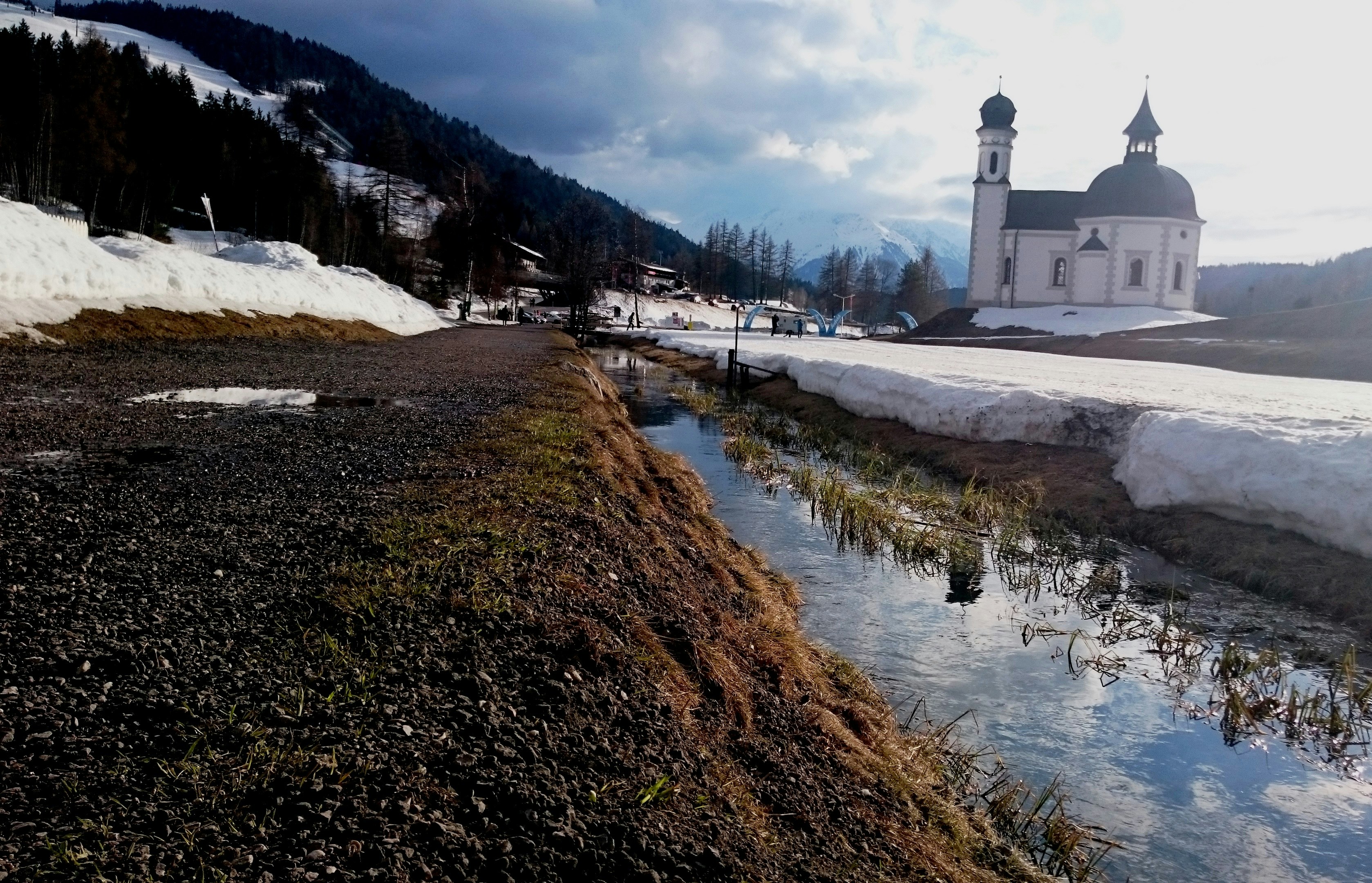 Riverside path beside a melting snowbank leads toward a small chapel with domed towers, reflecting in a narrow stream that runs beside the gravel road.