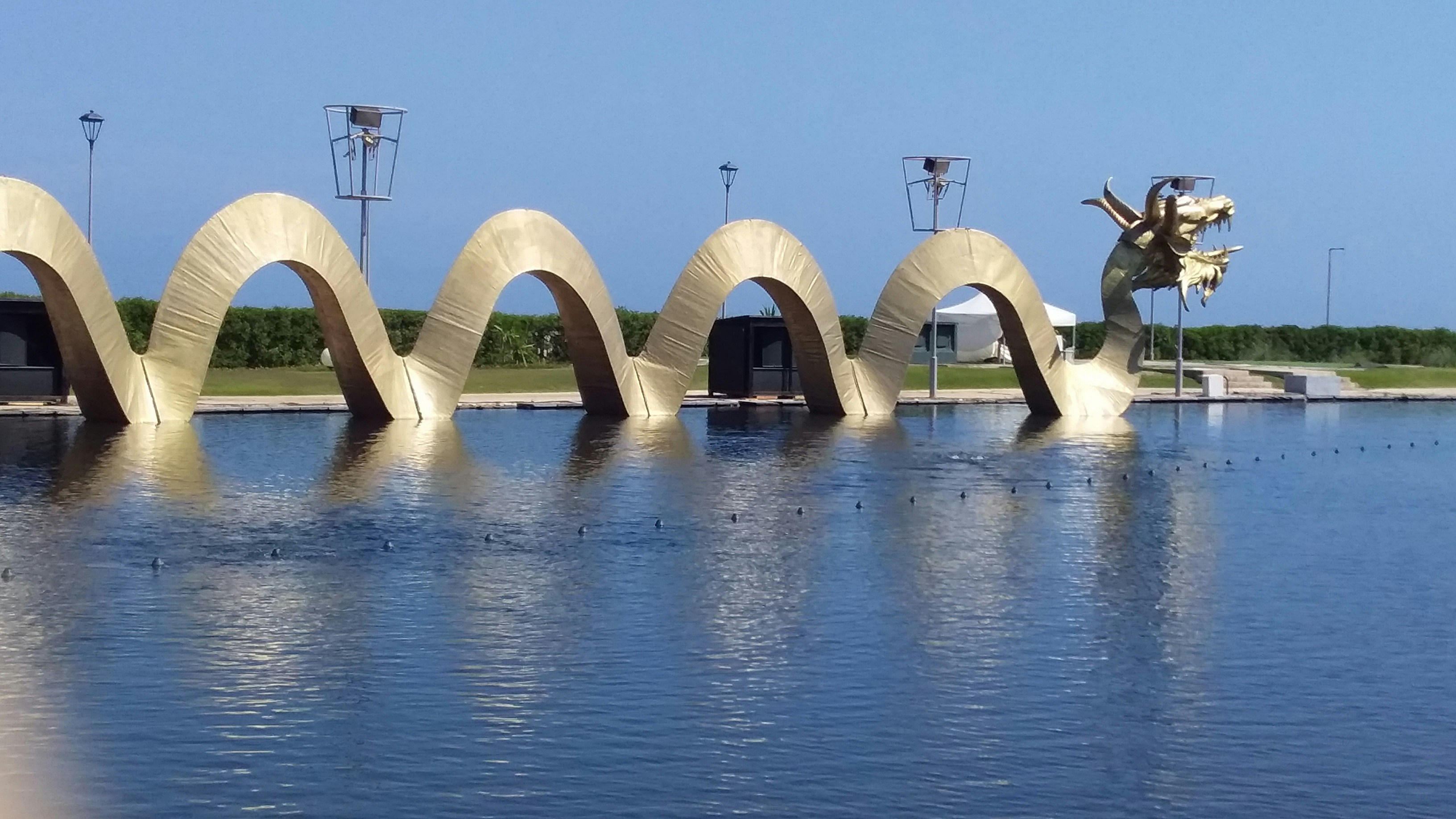 A striking golden sculpture of a dragon emerges from a serene body of water, surrounded by gracefully arched structures. The scene reflects a harmonious blend of art and nature.