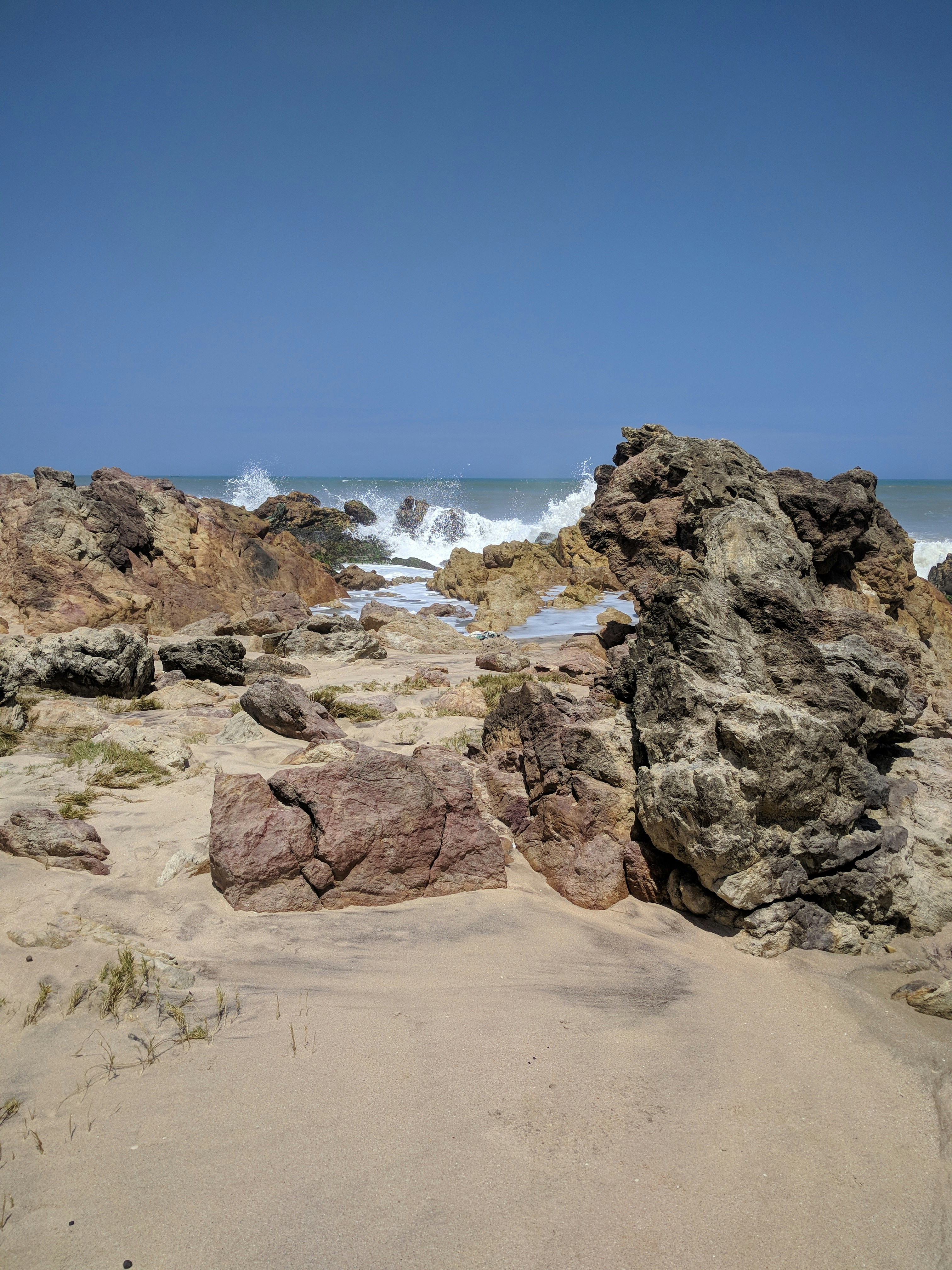 Rugged rocks frame a tranquil beach scene as gentle waves lap at the shore under a clear blue sky.