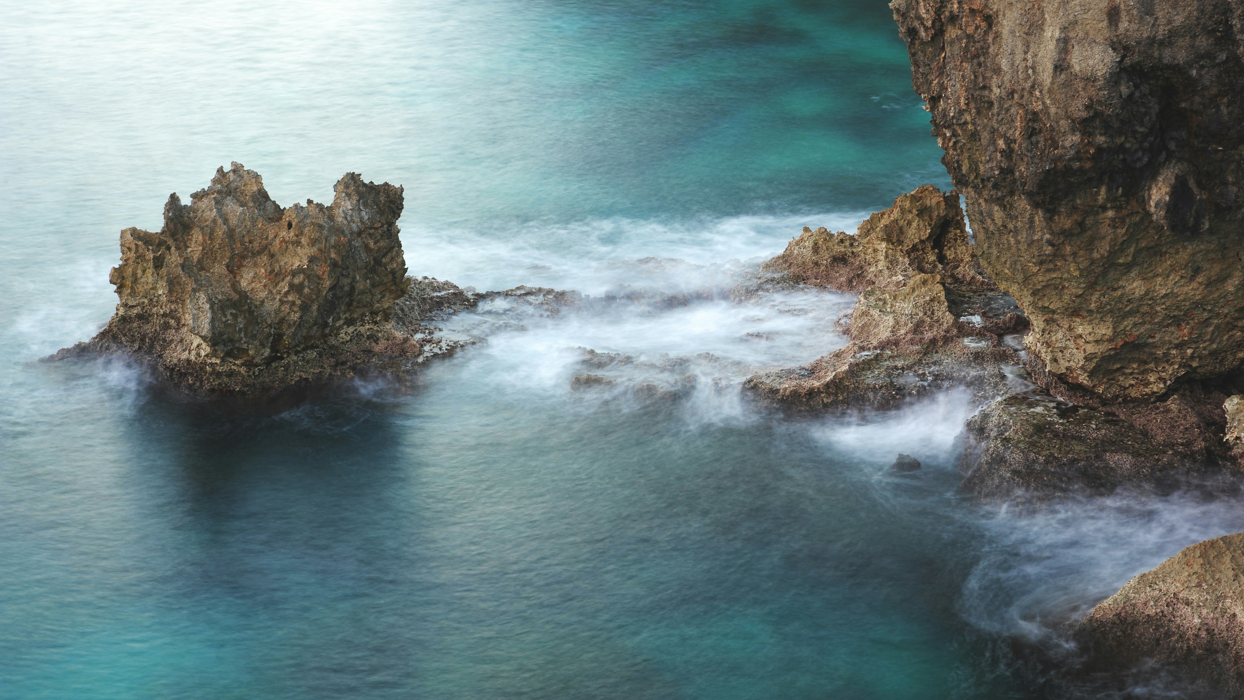 Rugged coastal rocks emerge from tranquil turquoise waters, with gentle waves lapping at their bases. The scene captures the serene interaction between land and sea.