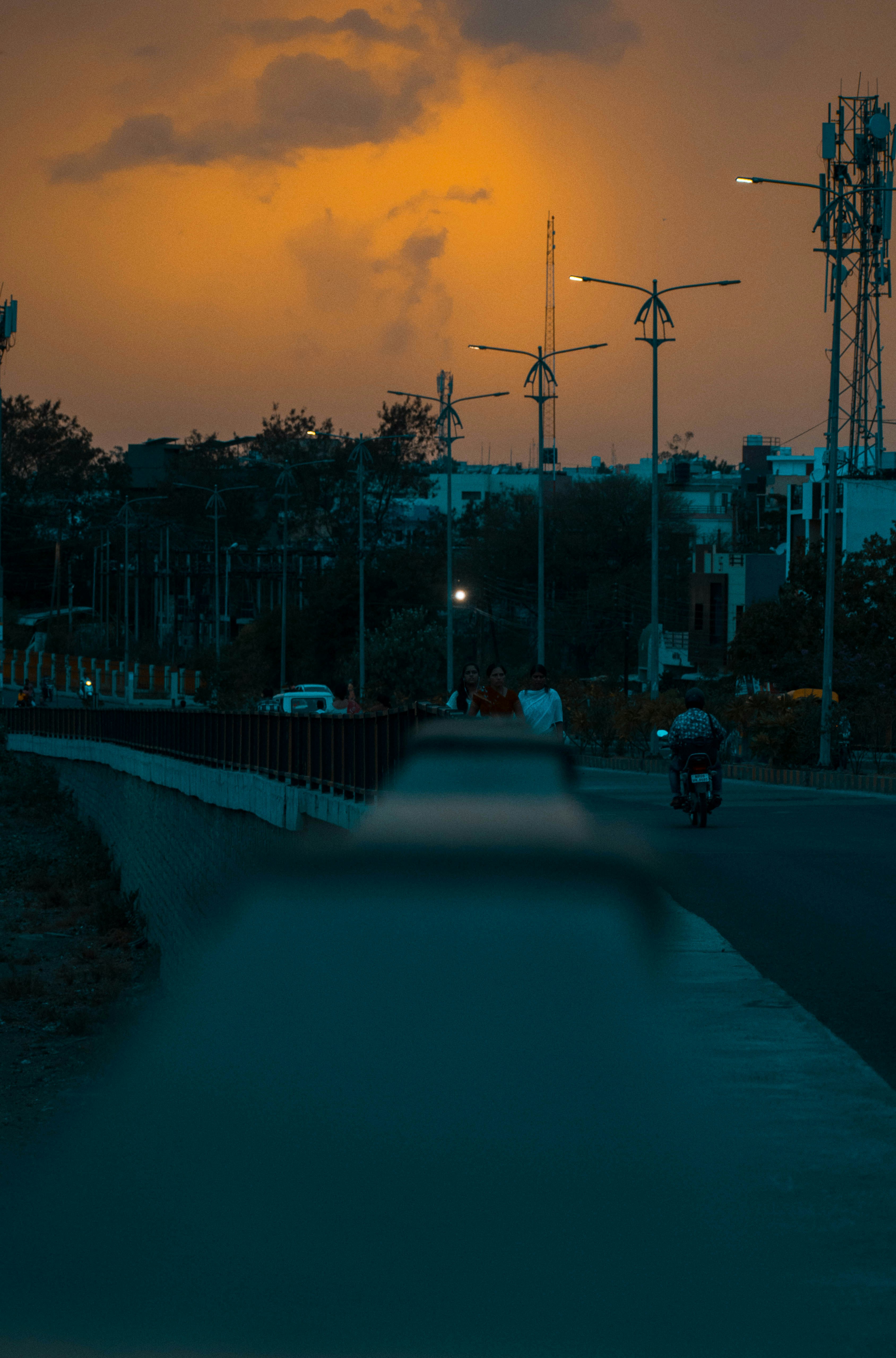 Silhouetted figures stroll along a city road under a glowing twilight sky, with streetlights illuminating the scene. A motorcycle rides past, adding motion to the tranquil urban landscape.