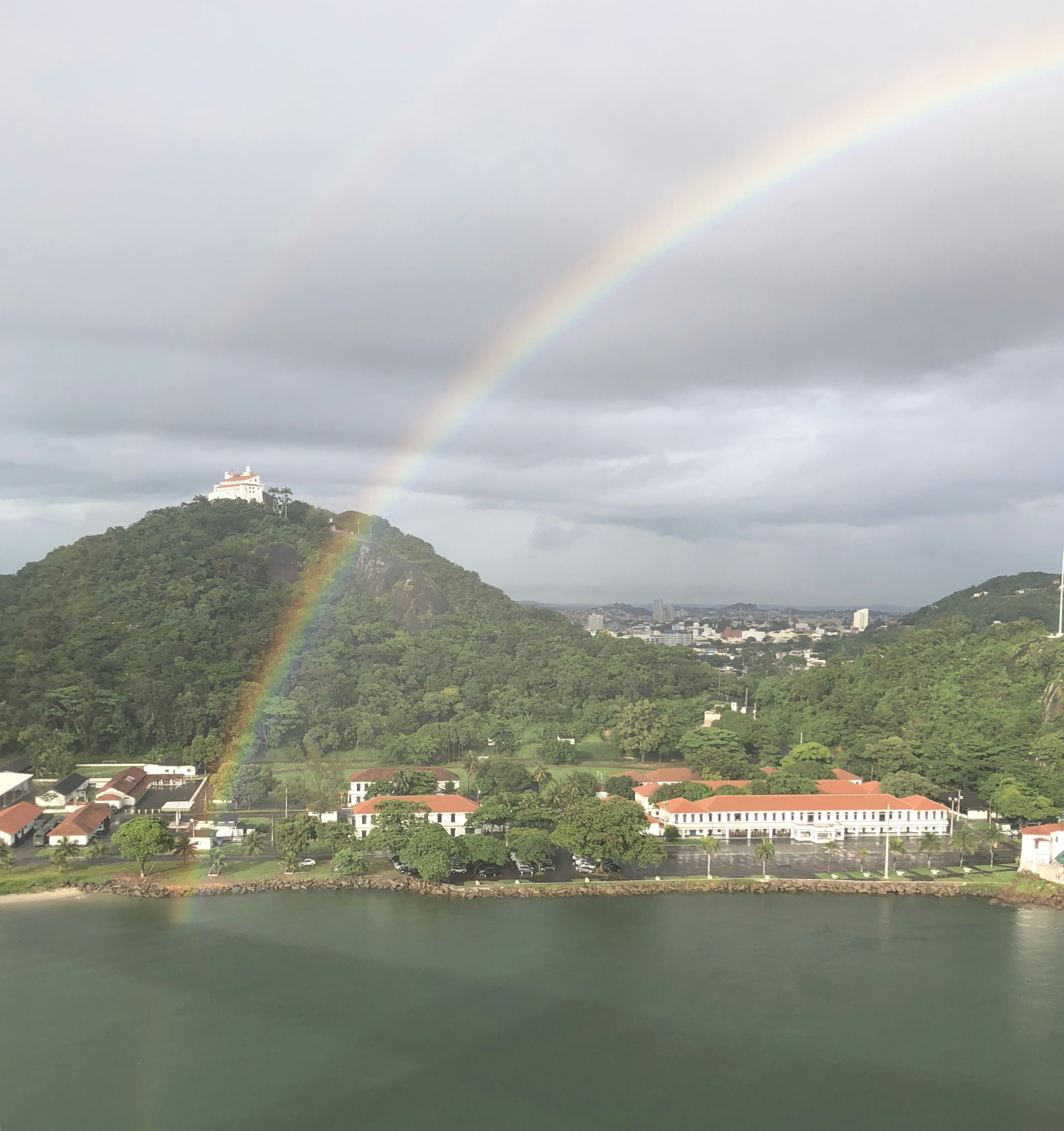 Vibrant rainbow arching over lush hills and a coastal town, blending nature with urban elements. The scene captures a tranquil moment after rain.