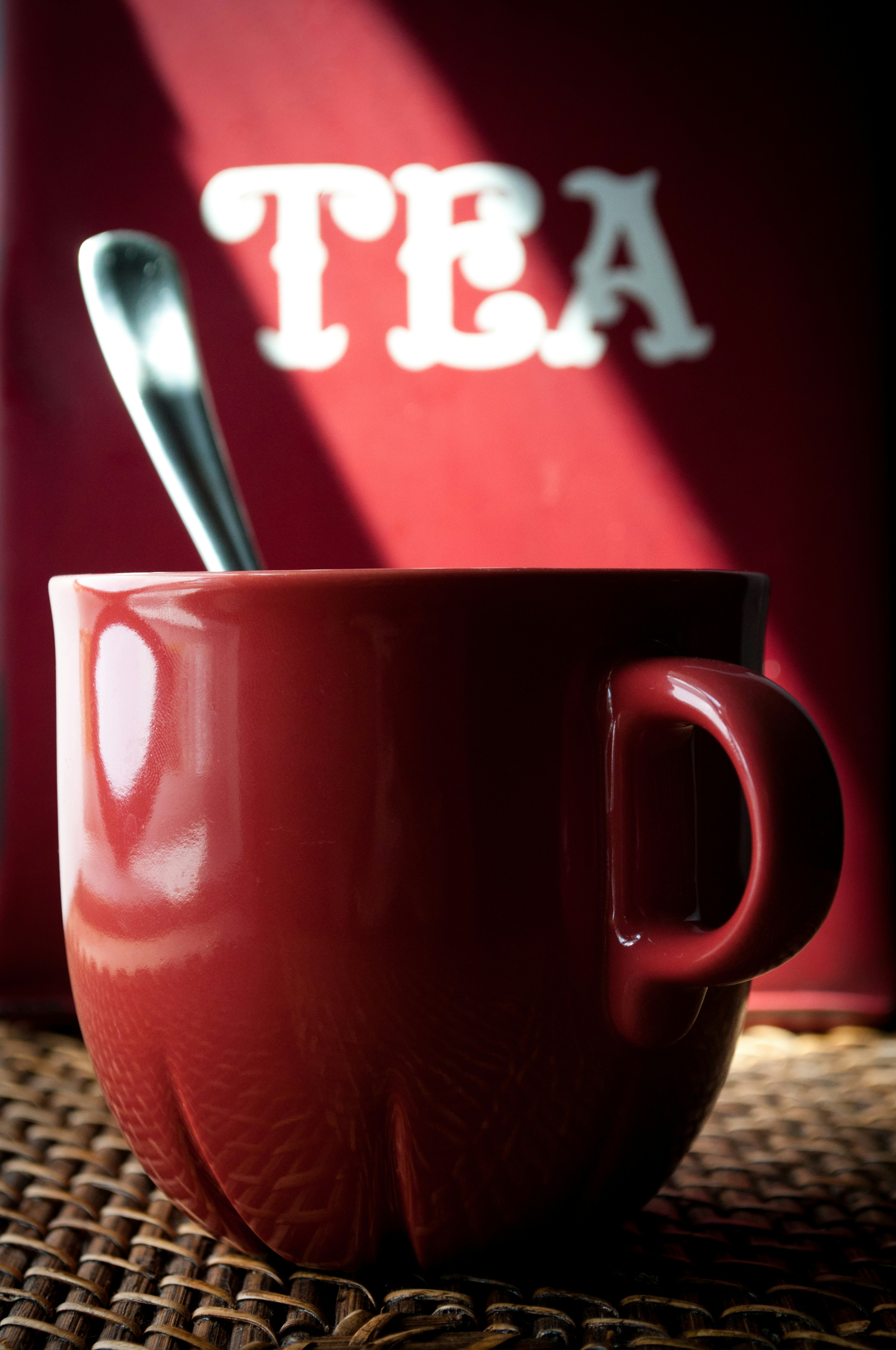 Red ceramic mug filled with tea, featuring a silver spoon resting inside, set against a backdrop with the word 'TEA' in bold letters. 