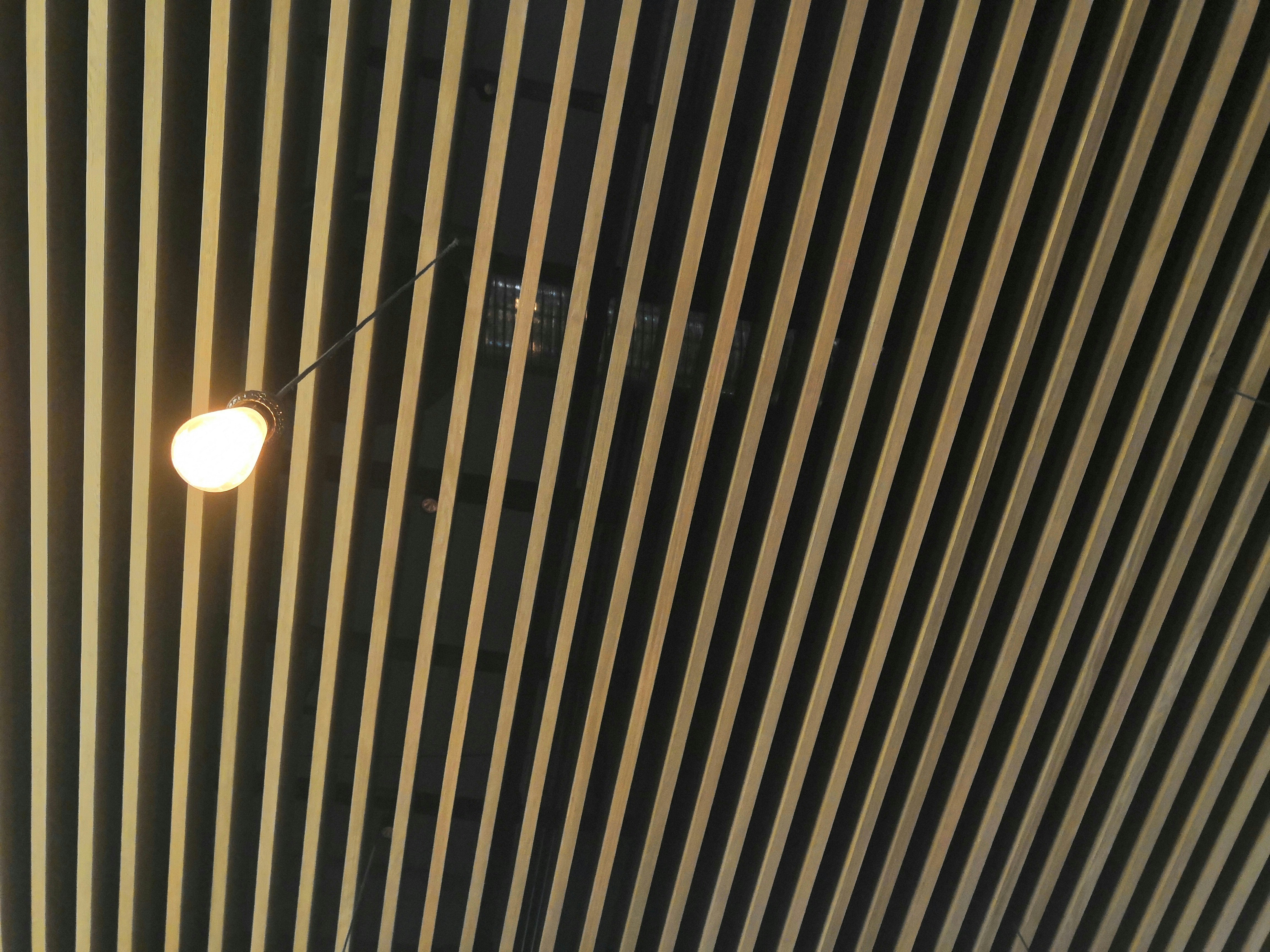 Photograph of a ribbed ceiling with a single warm light bulb casting a soft glow. The vertical slats create bold lines that guide the eye through the frame.
