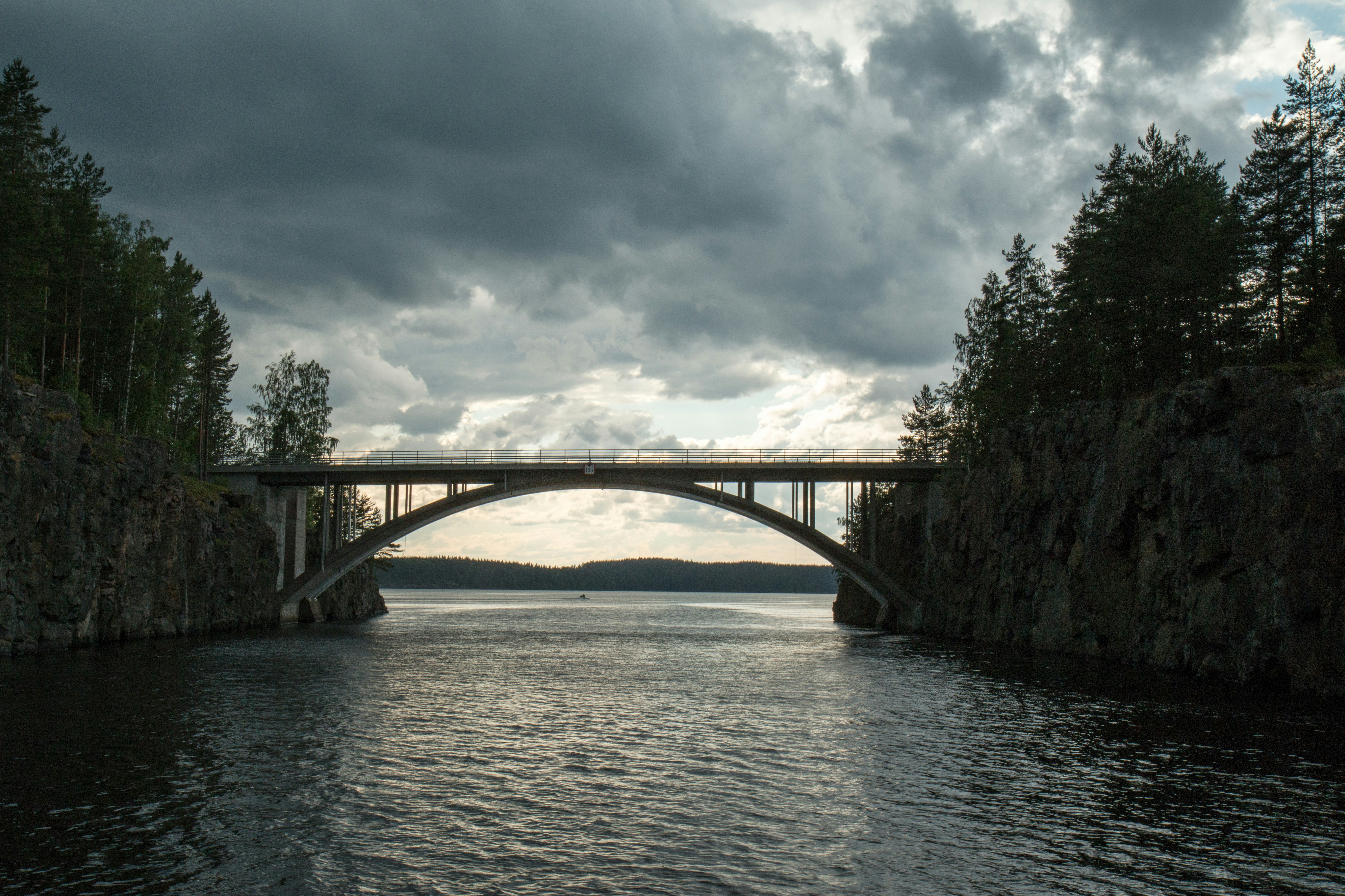 A modern bridge arches over a serene body of water, framed by rocky cliffs and dense forest under a cloudy sky.