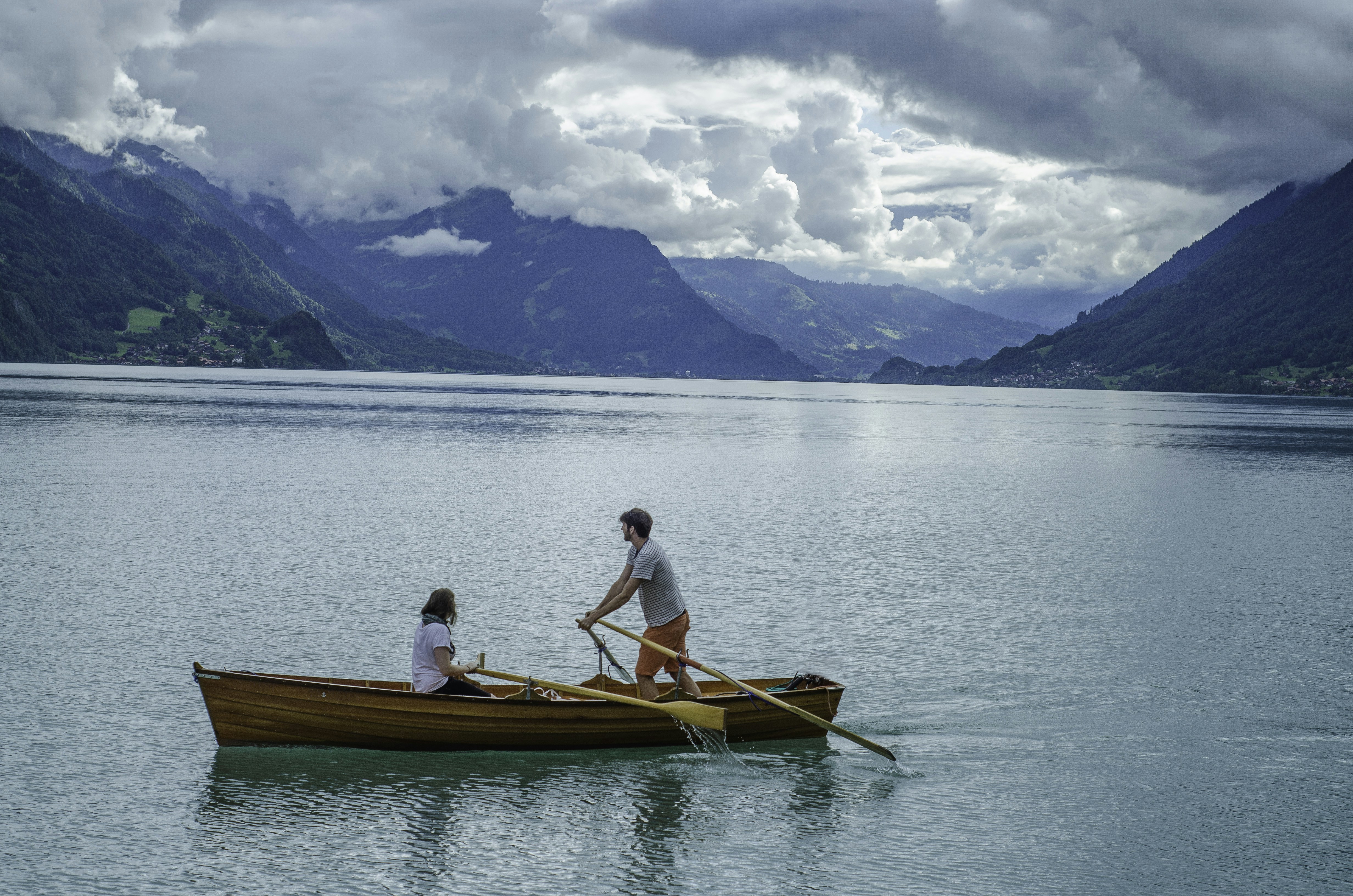 Two individuals in a wooden rowboat glide across a tranquil lake surrounded by majestic mountains and dramatic clouds.