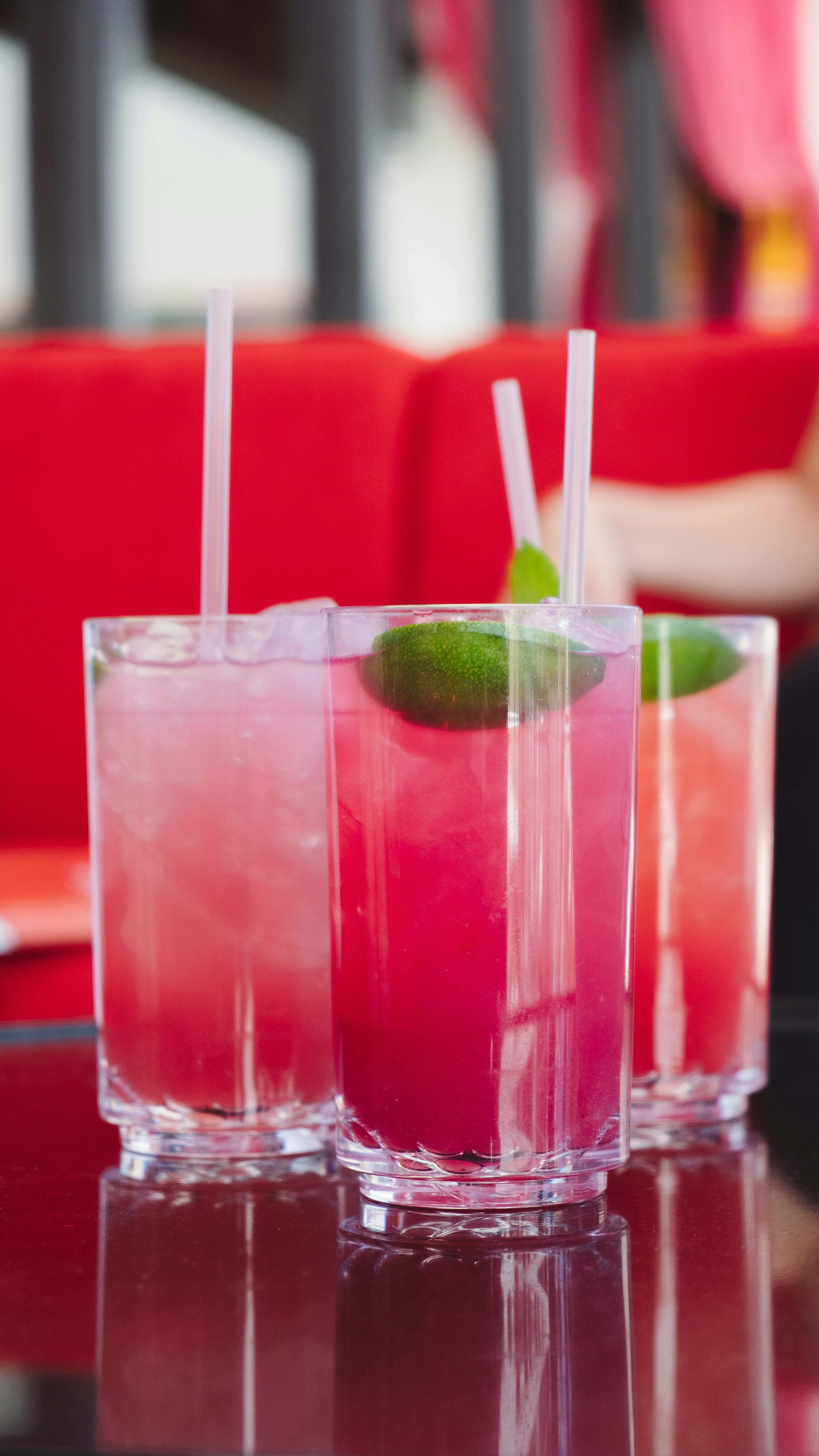 Three colorful cocktails with lime and mint garnishes sit on a table, reflecting vibrant hues against a red backdrop.