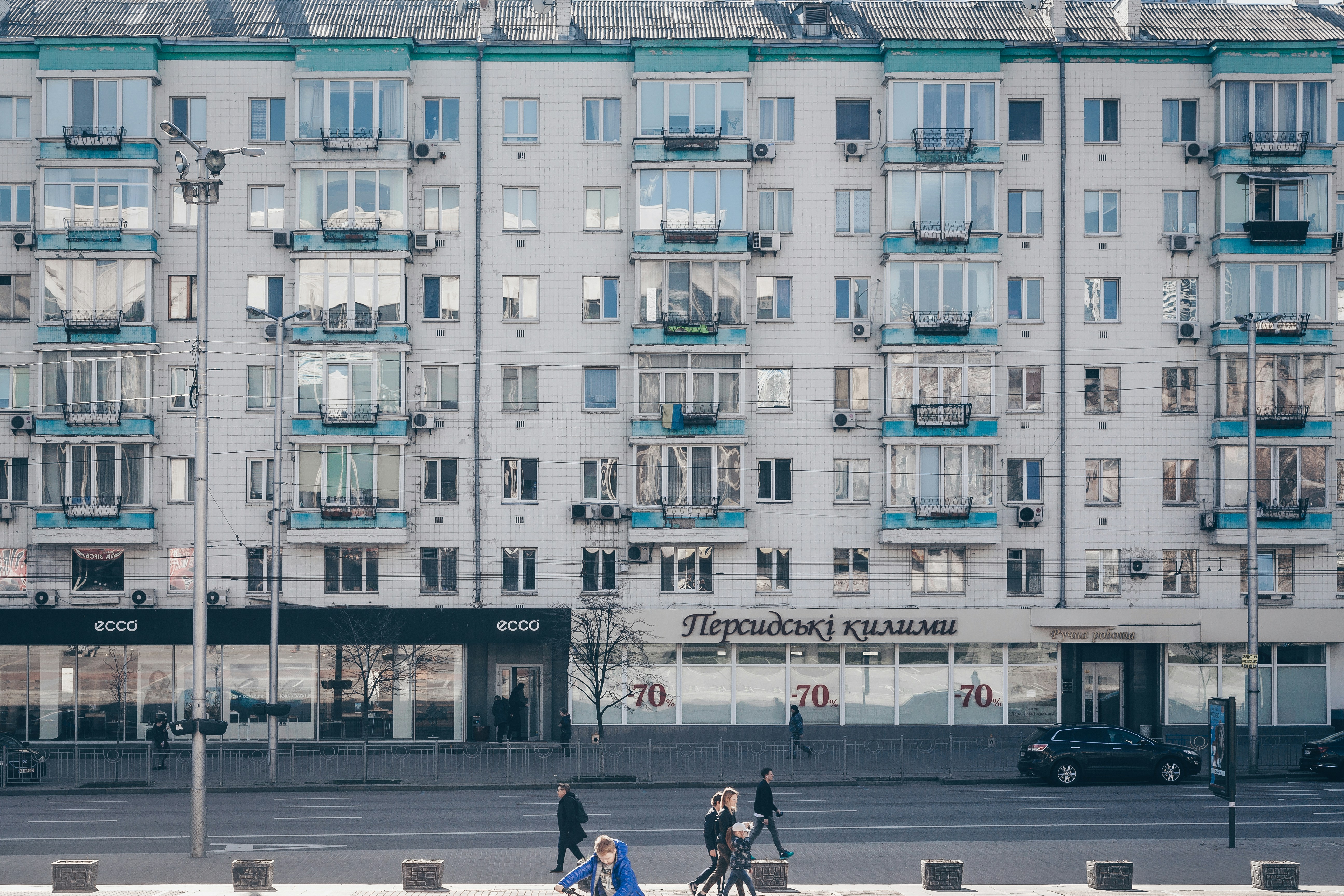 Pedestrians walk past a symmetrical facade of an apartment building with teal balconies and shop signs below.