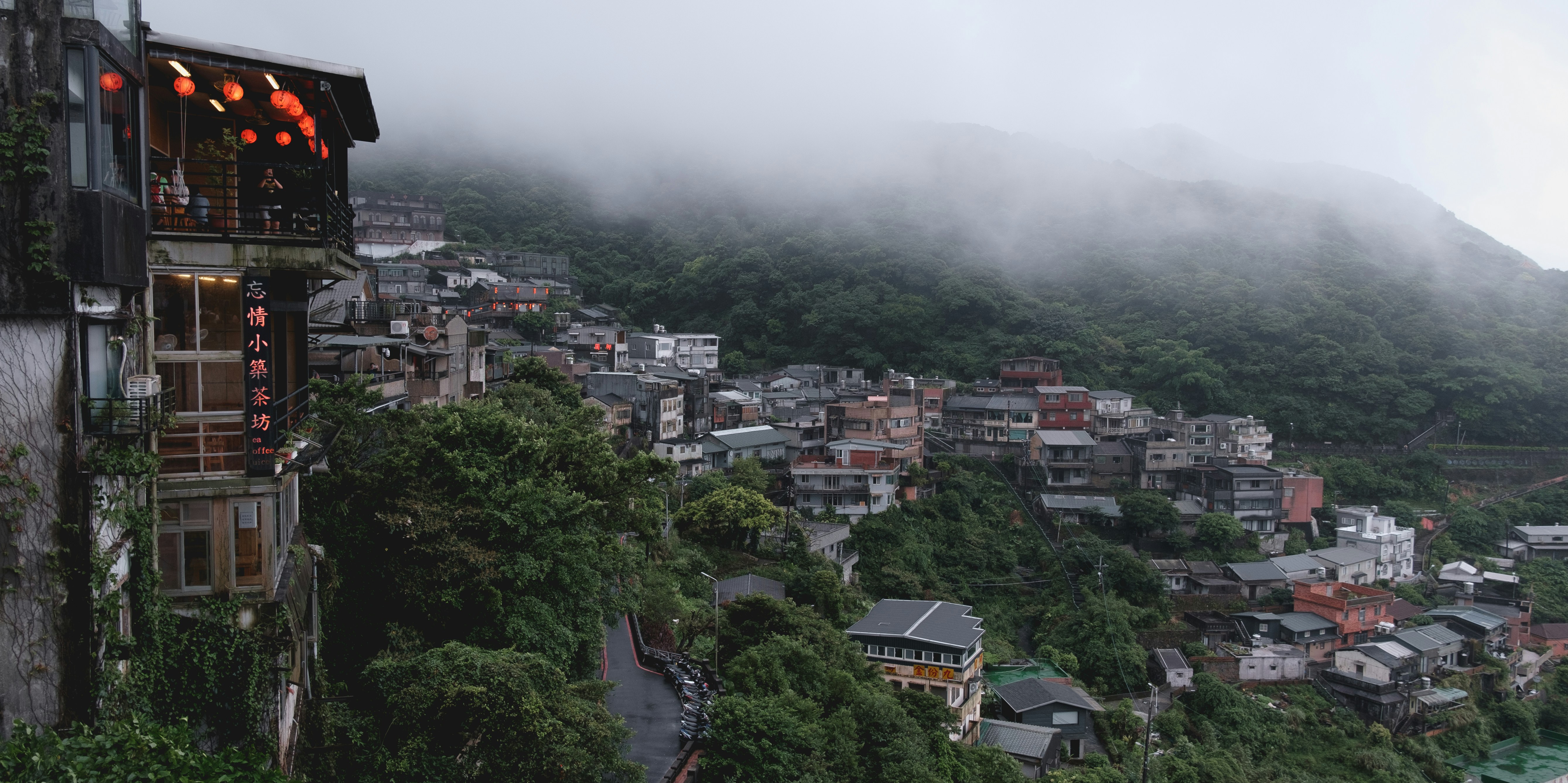 A hillside village enveloped in mist, showcasing traditional architecture and vibrant lanterns. The lush greenery contrasts with the quaint buildings nestled against the mountainside.