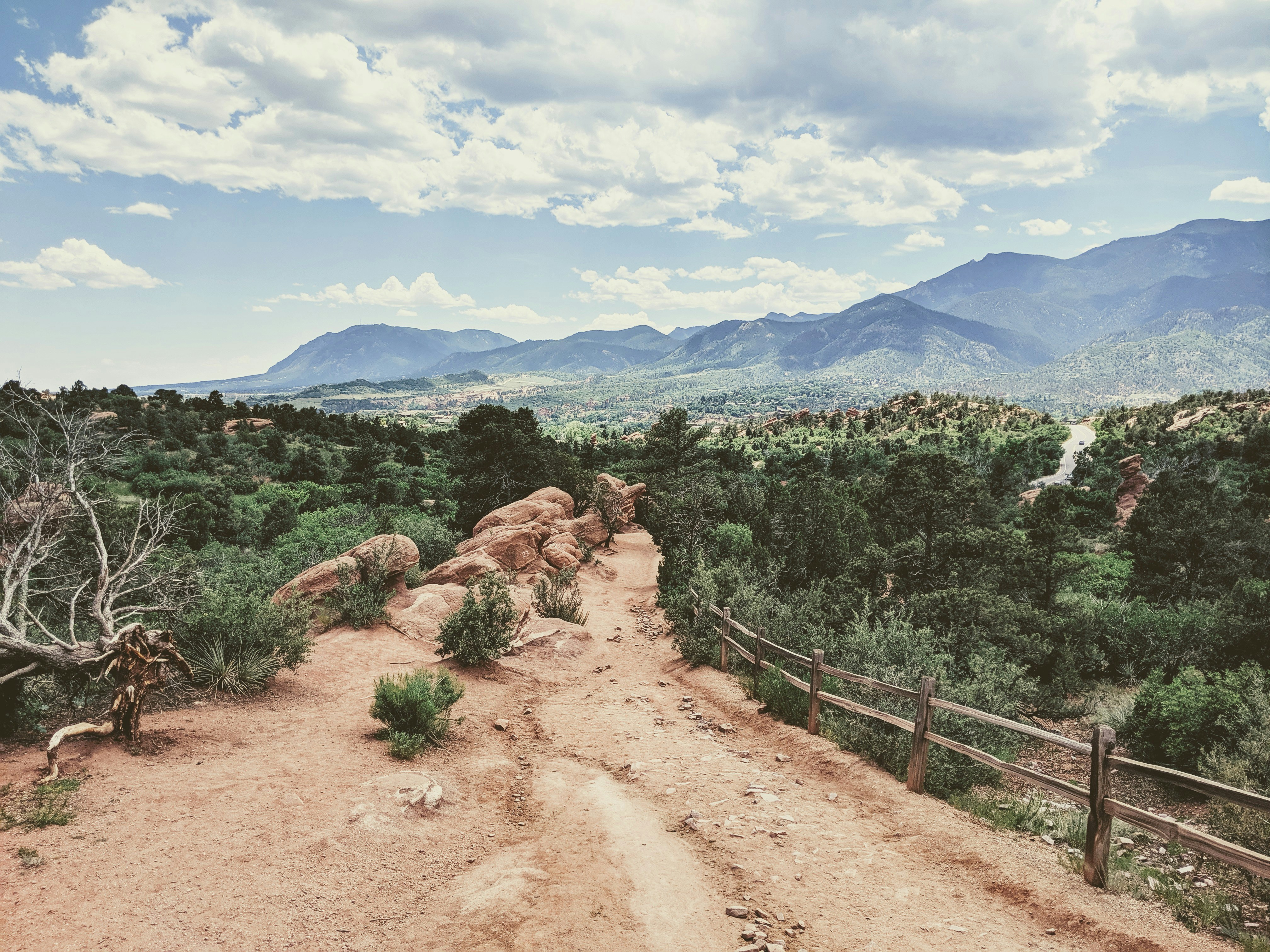 Dusty trail bordered by a wooden fence winds through a rugged landscape with distant mountains under a partly cloudy sky.