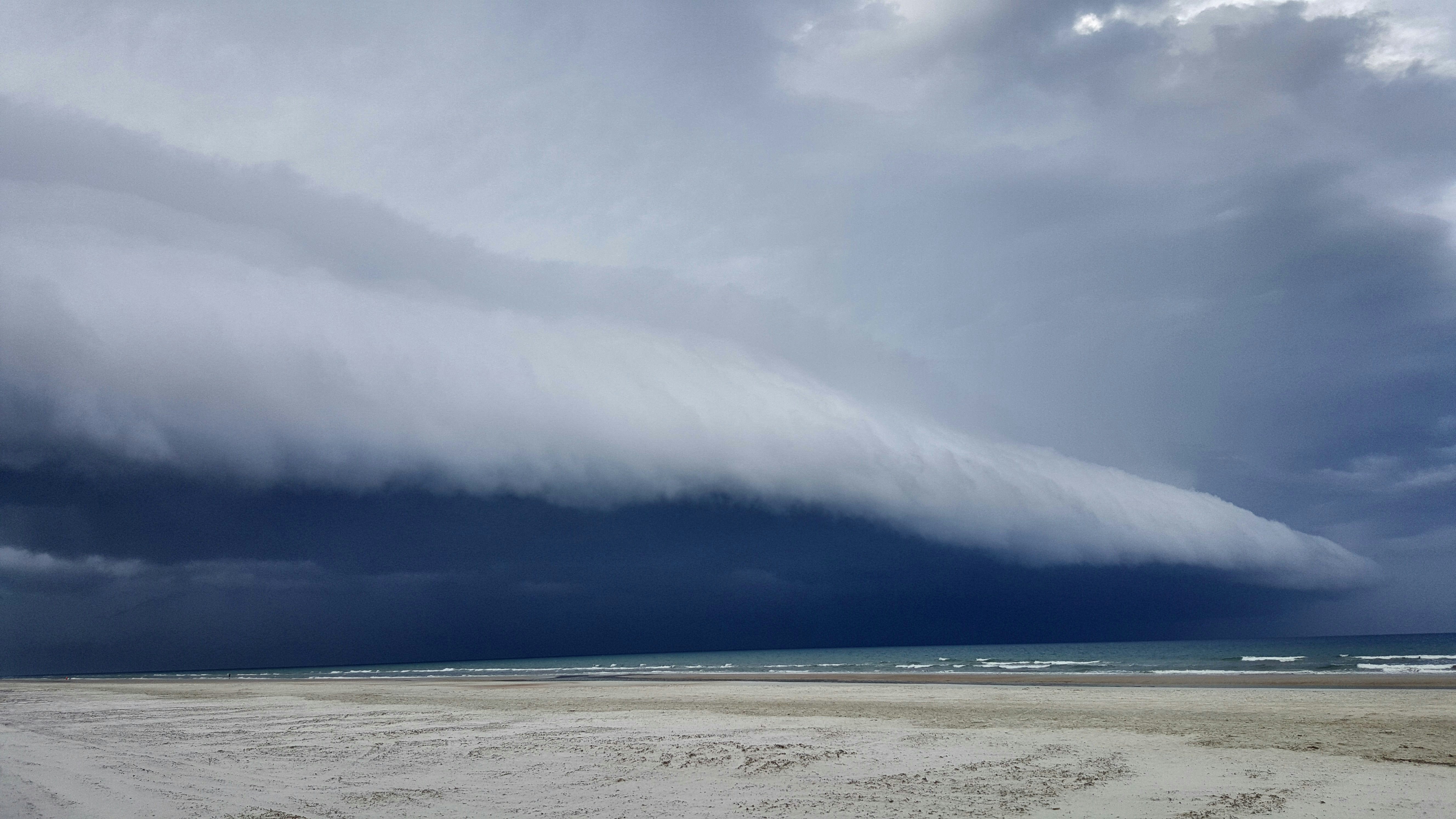 Massive cloud formation stretches across a tranquil beach with waves gently lapping the shore.