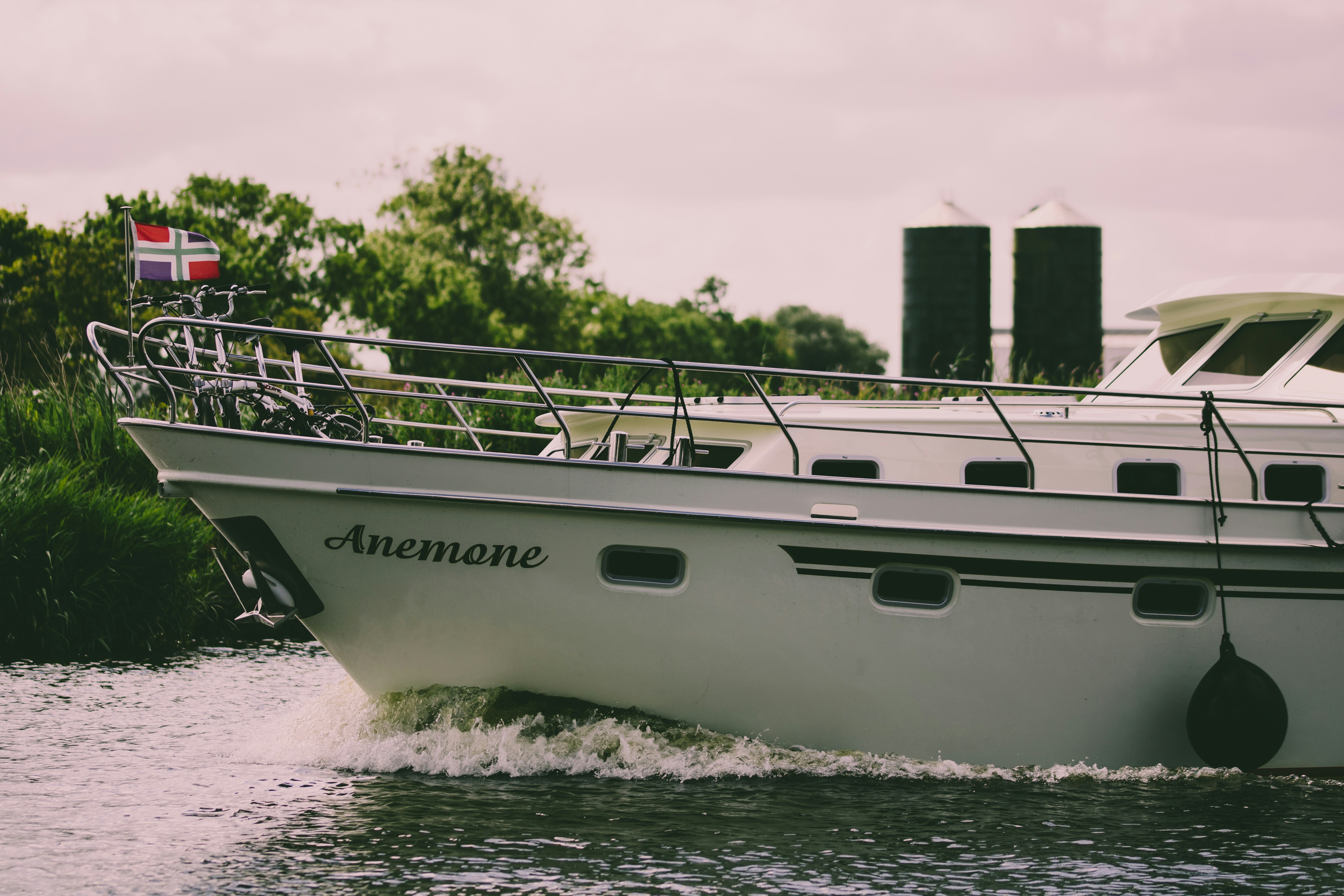 White boat named Anemone gliding through calm waters, flanked by lush greenery and distant silos.