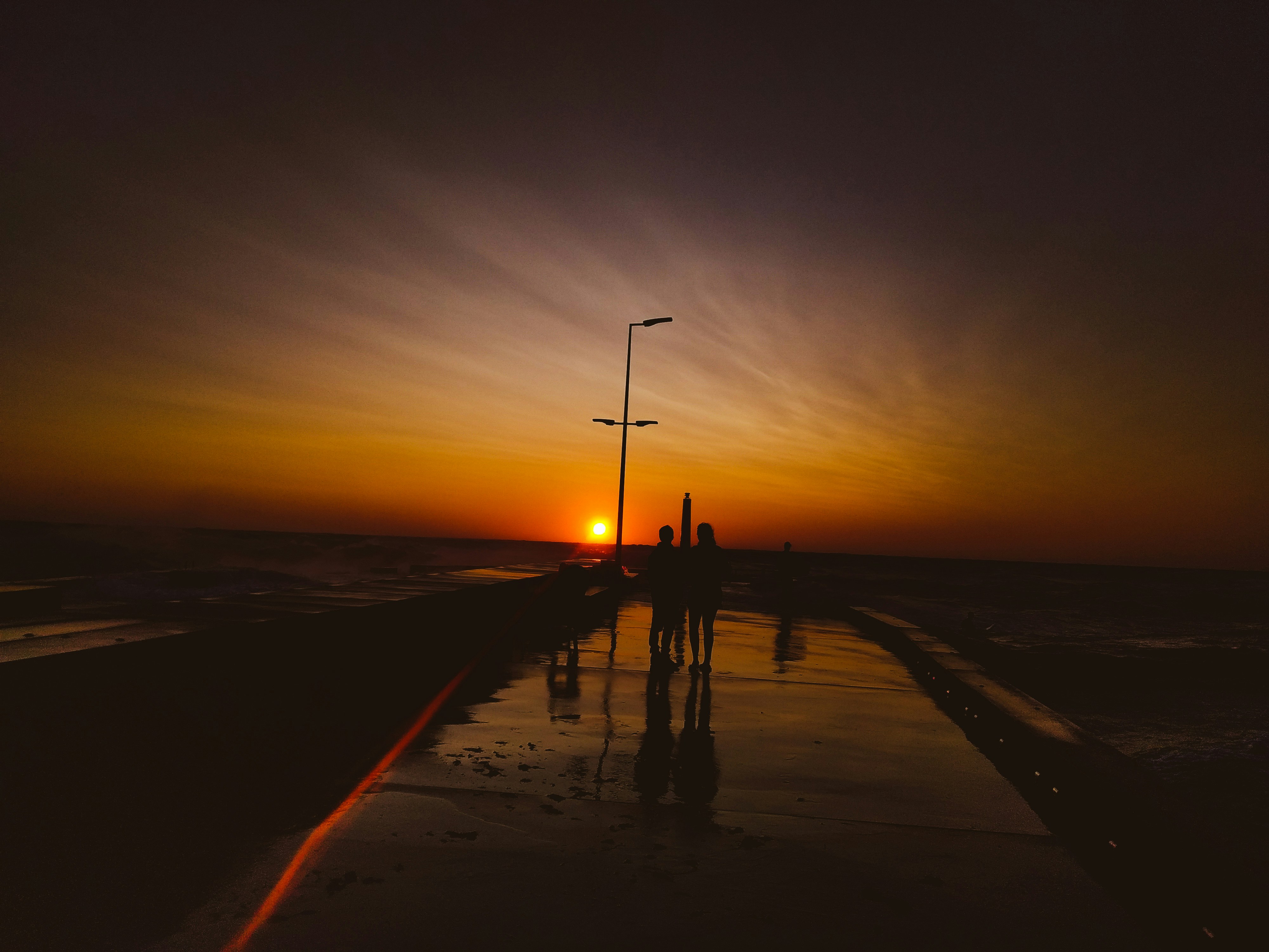 Silhouetted figures walking hand in hand on a wet promenade at sunset, with streetlights illuminating the scene against a vibrant sky.