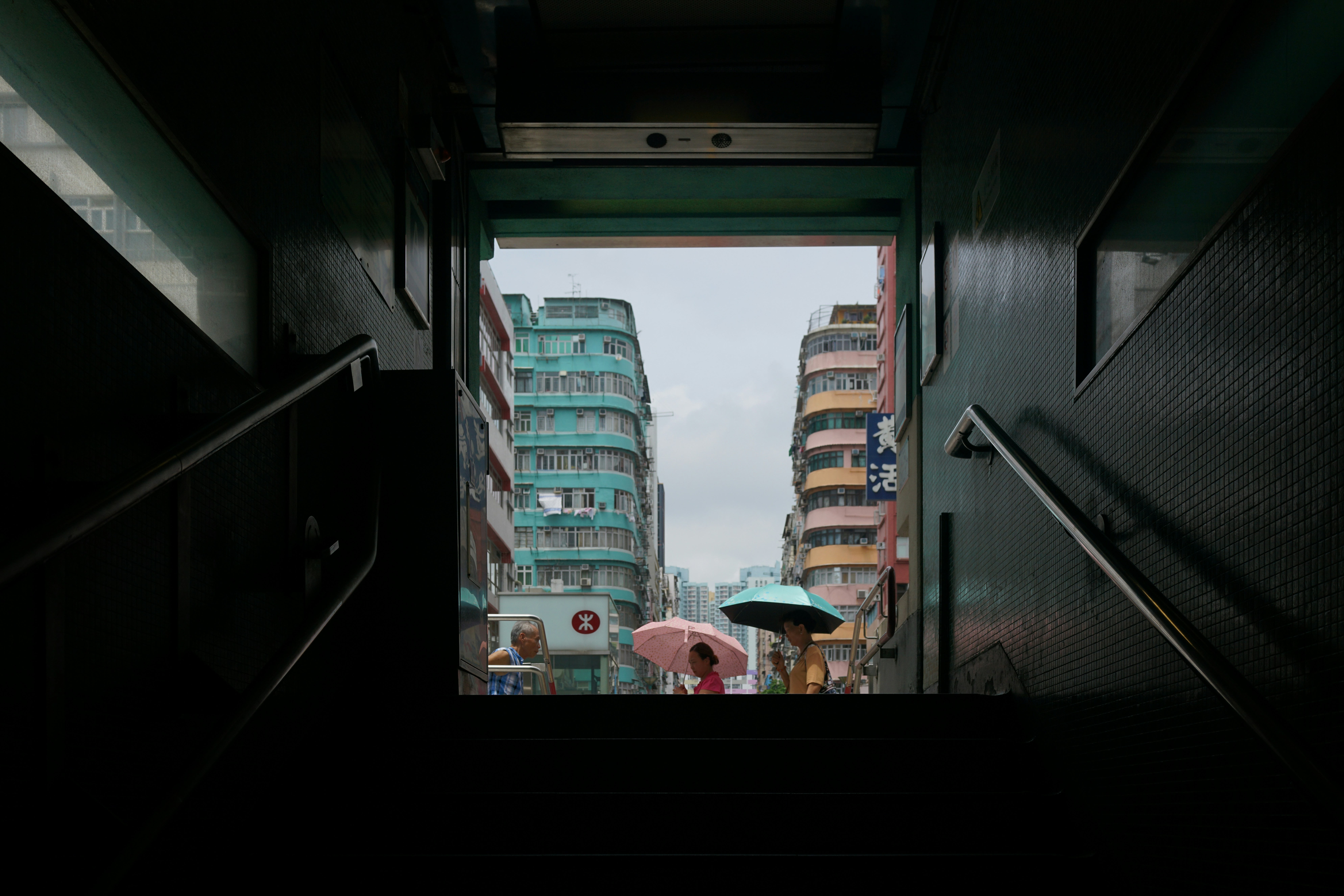 View from an underpass revealing a bustling city street with pedestrians holding umbrellas. The architectural lines and colors create a vibrant urban scene.