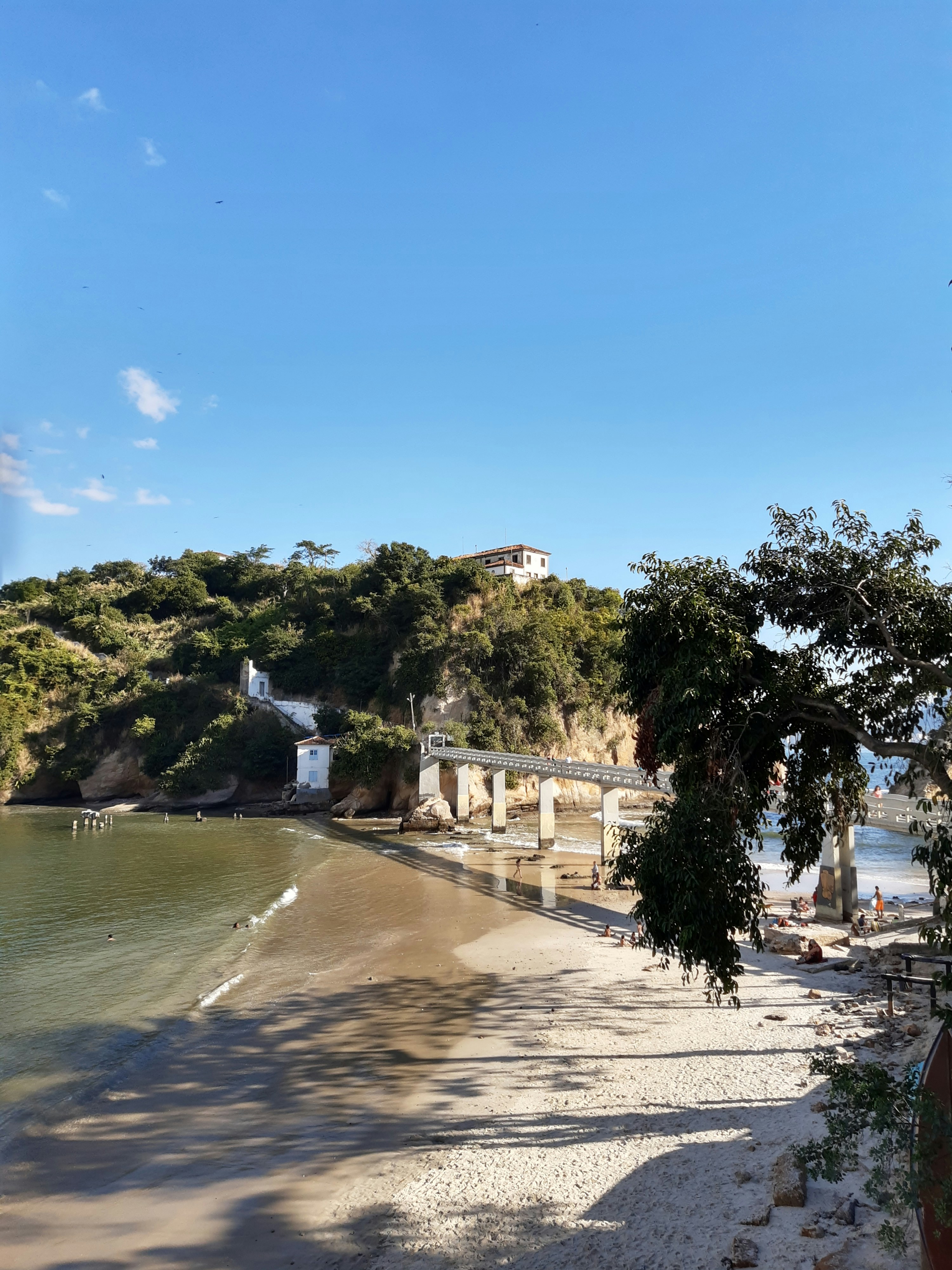 A scenic beach view featuring a bridge connecting the shoreline to a hillside, with people enjoying the water in the foreground.