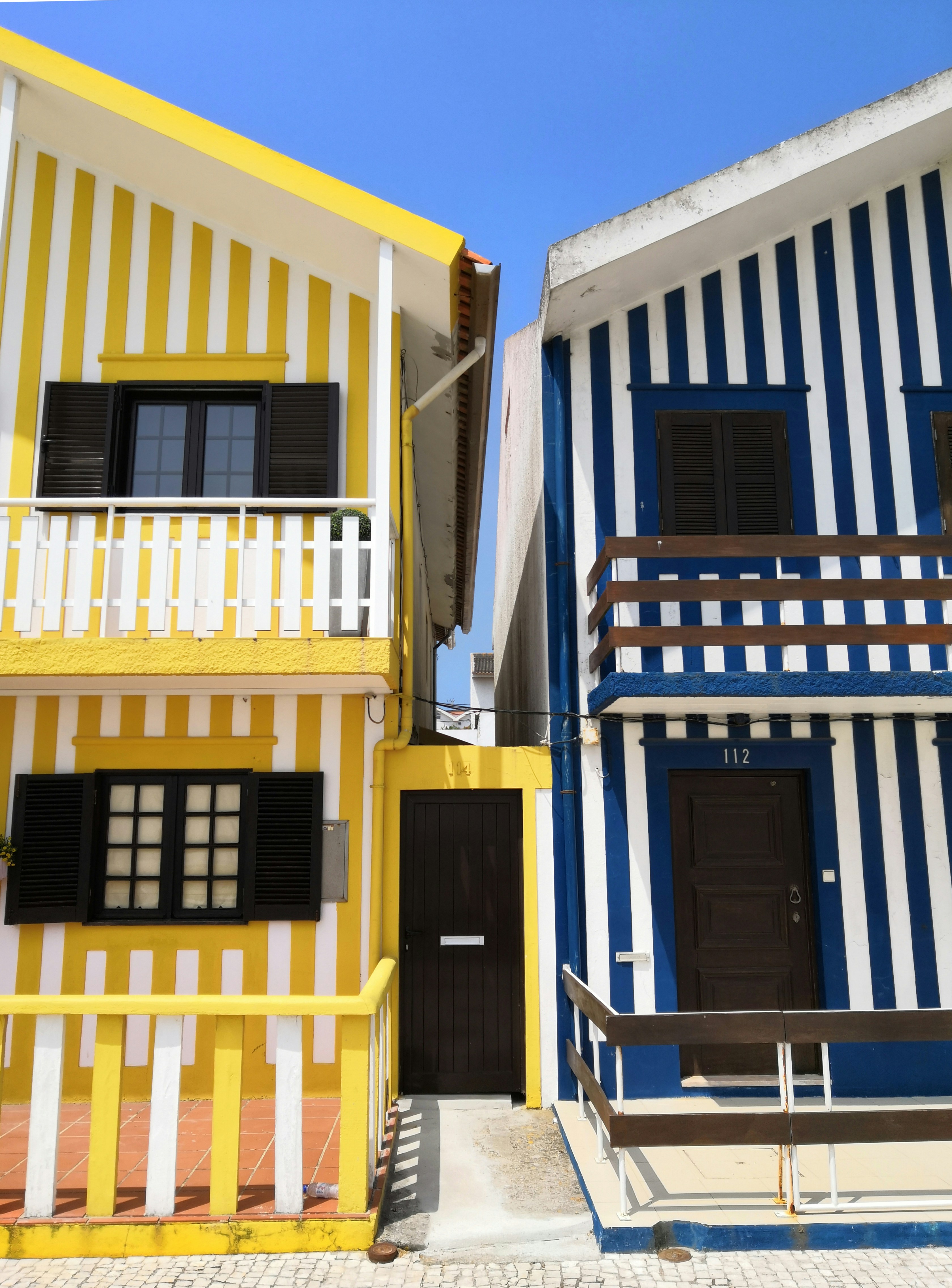 Two neighboring houses showcase bold yellow and blue vertical stripes that frame a narrow central doorway. The scene emphasizes color geometry against a clear blue sky.