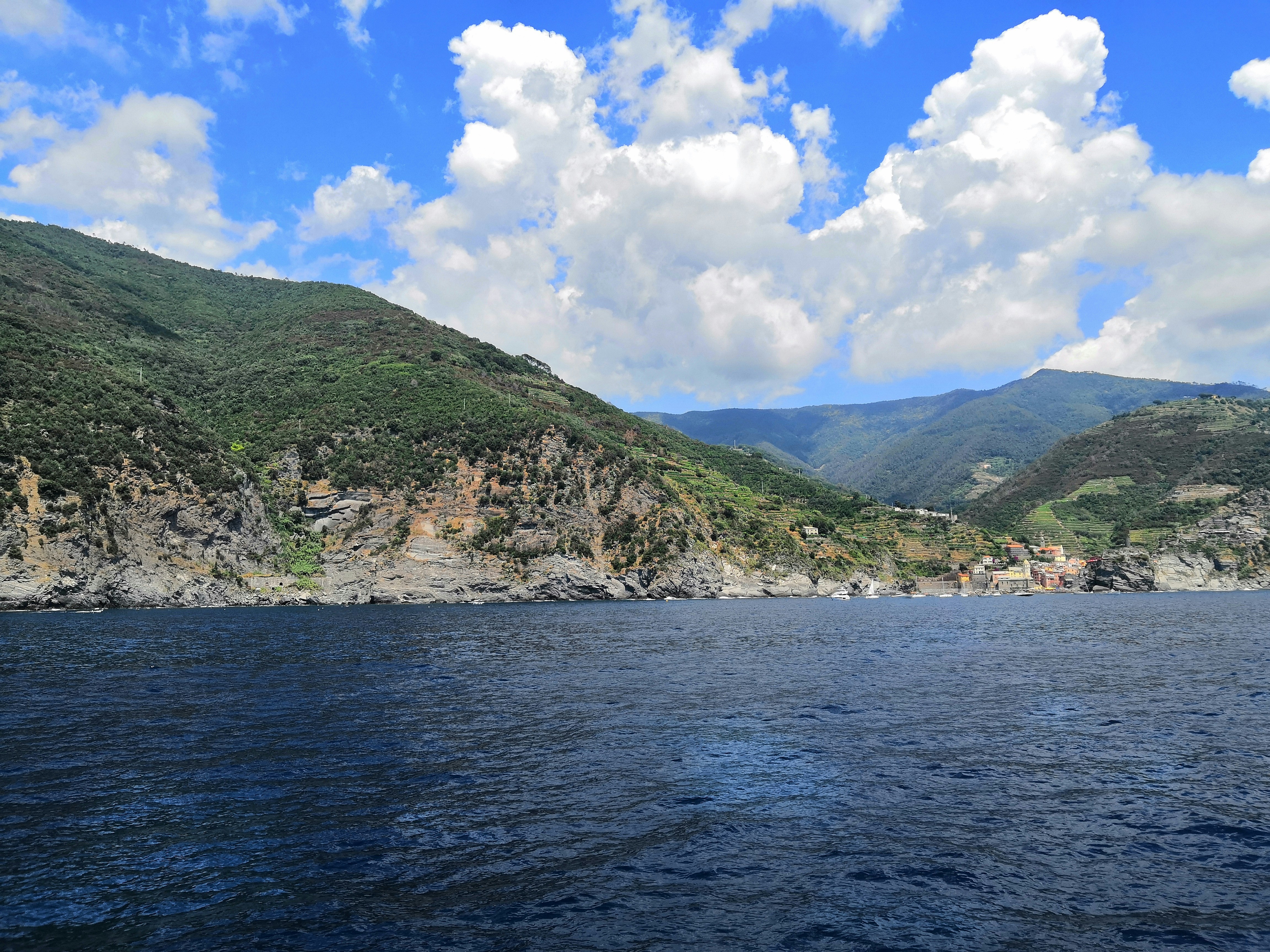 Coastal village tucked along rugged cliffs with calm blue sea in the foreground. A bright, cloud-filled sky crowns the landscape.