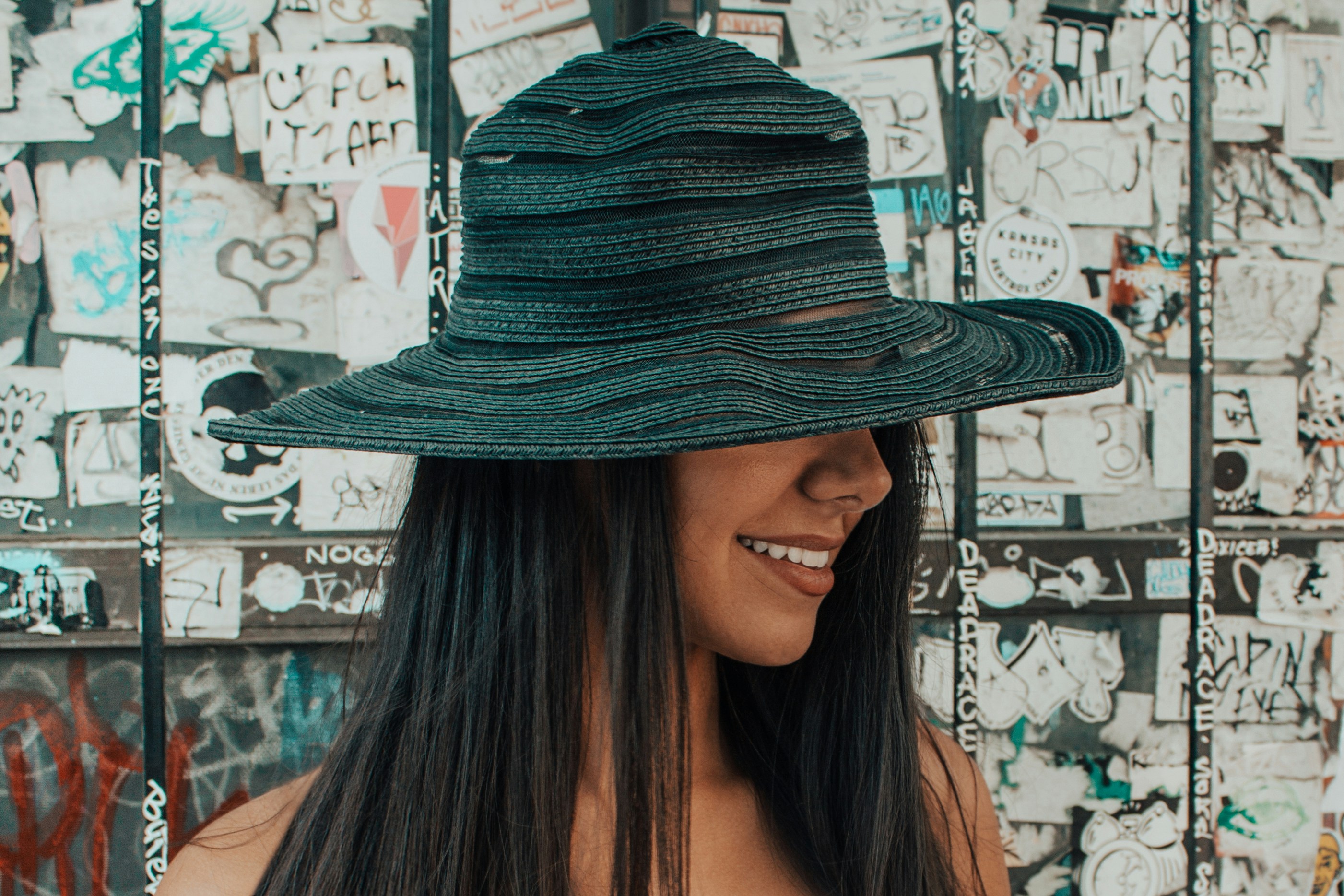 Woman wearing a wide-brimmed black hat, smiling softly against a backdrop of vibrant graffiti and stickers.