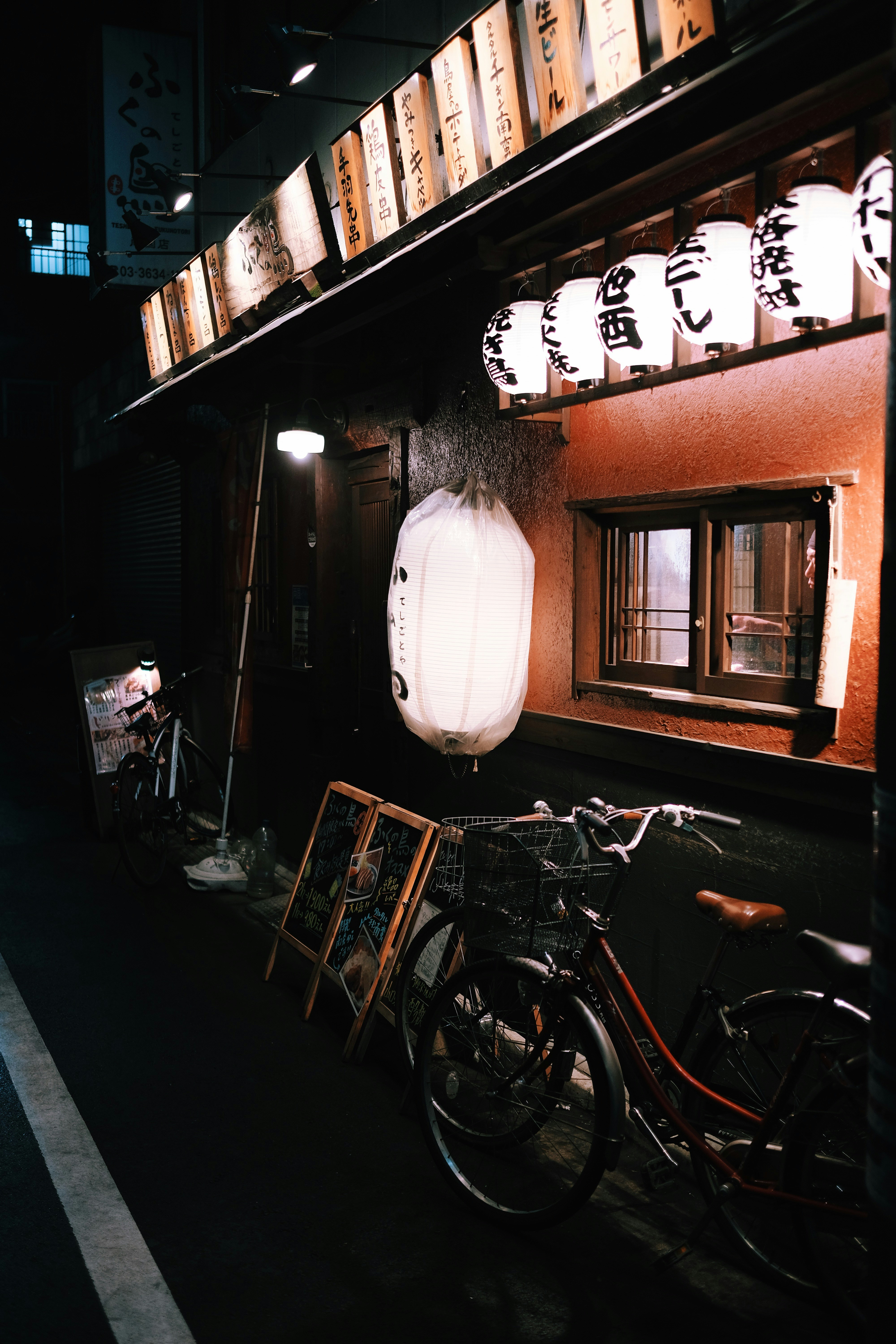 A traditional Japanese lantern illuminates a quaint street scene, highlighting bicycles and wooden signage in a cozy urban setting.