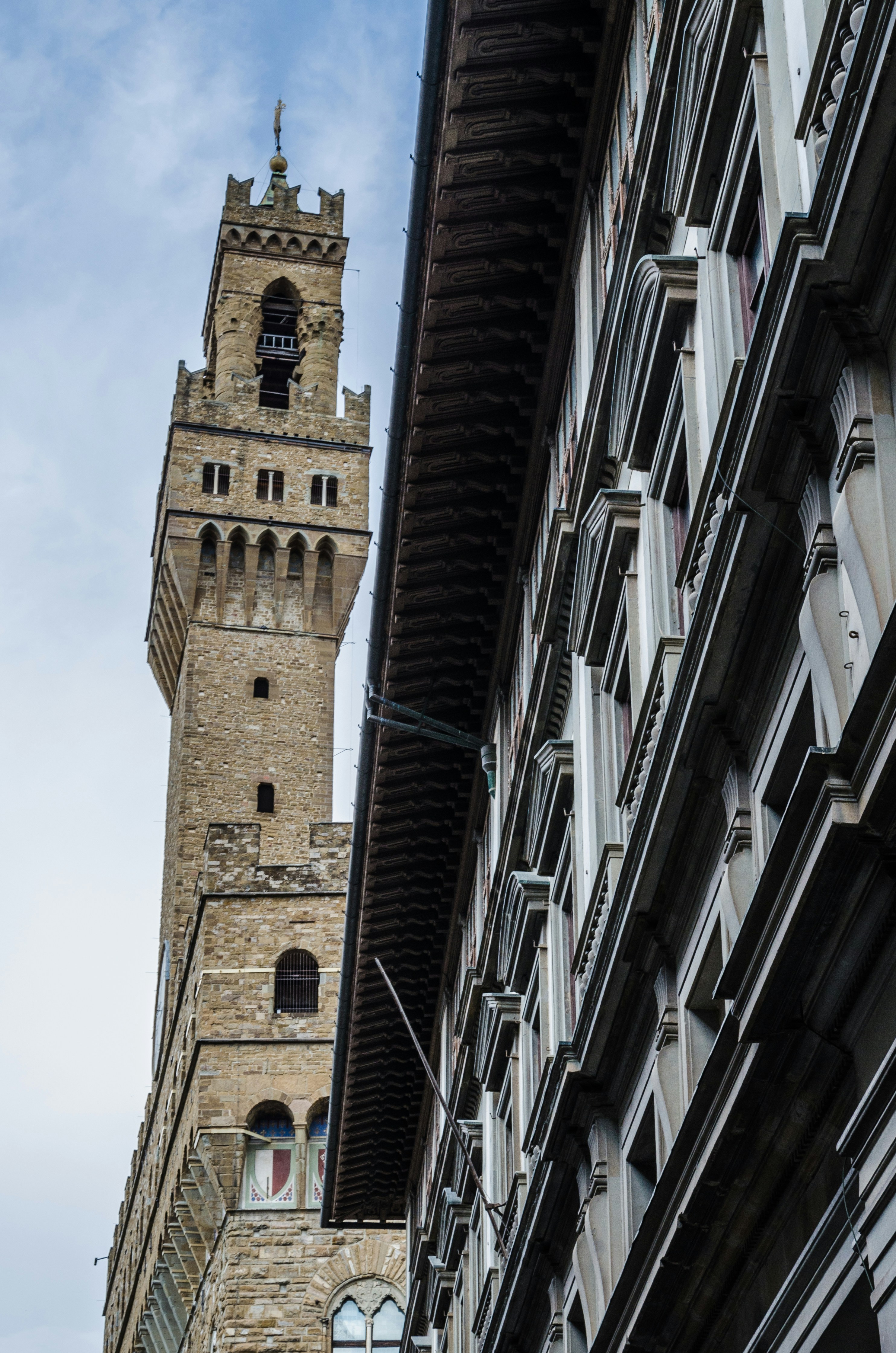Historic tower rising above the architectural details of a nearby building against a cloudy sky.