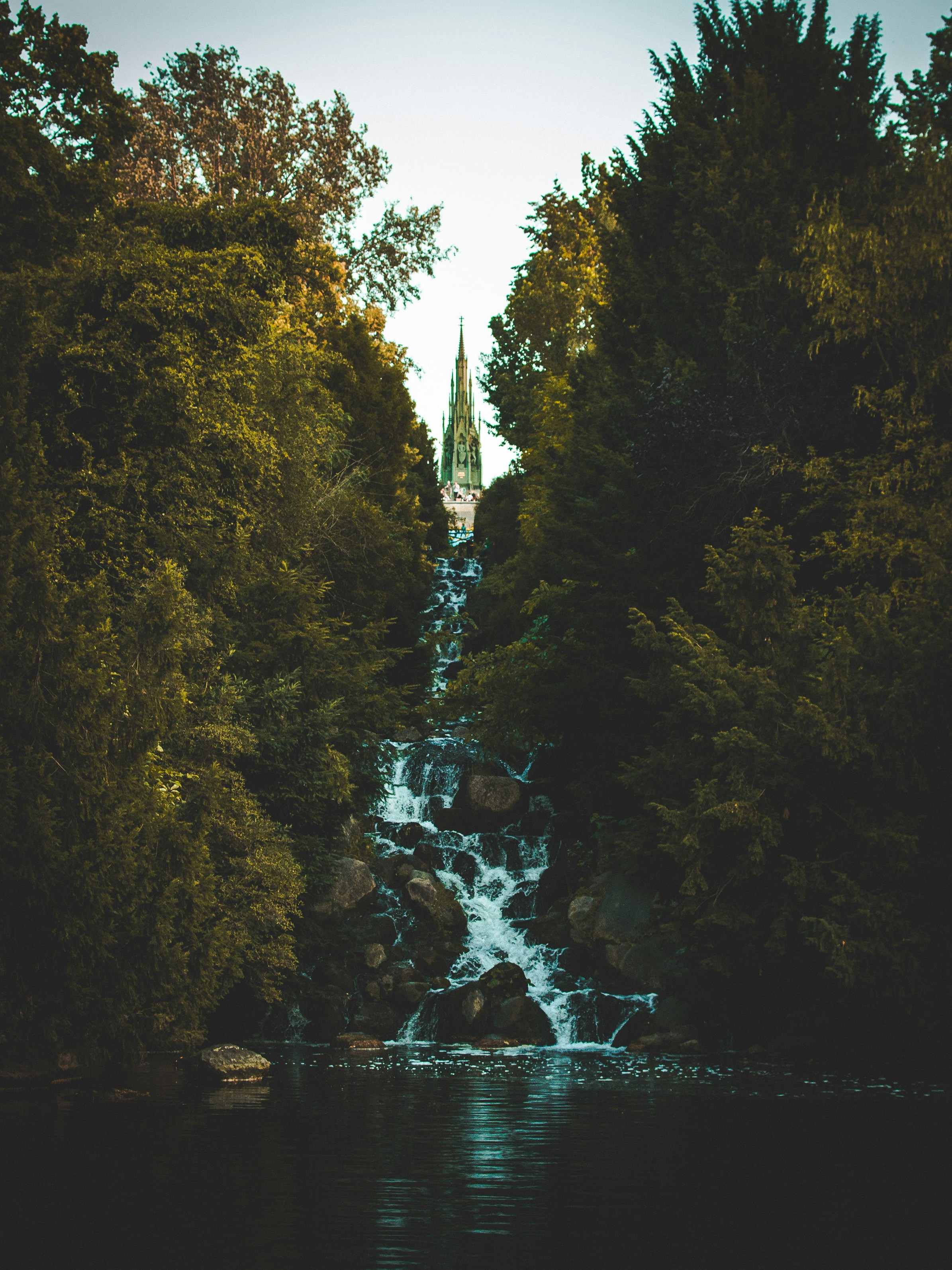 A tranquil waterfall flows through a lush green landscape, leading the eye toward a distant steeple rising above the trees.
